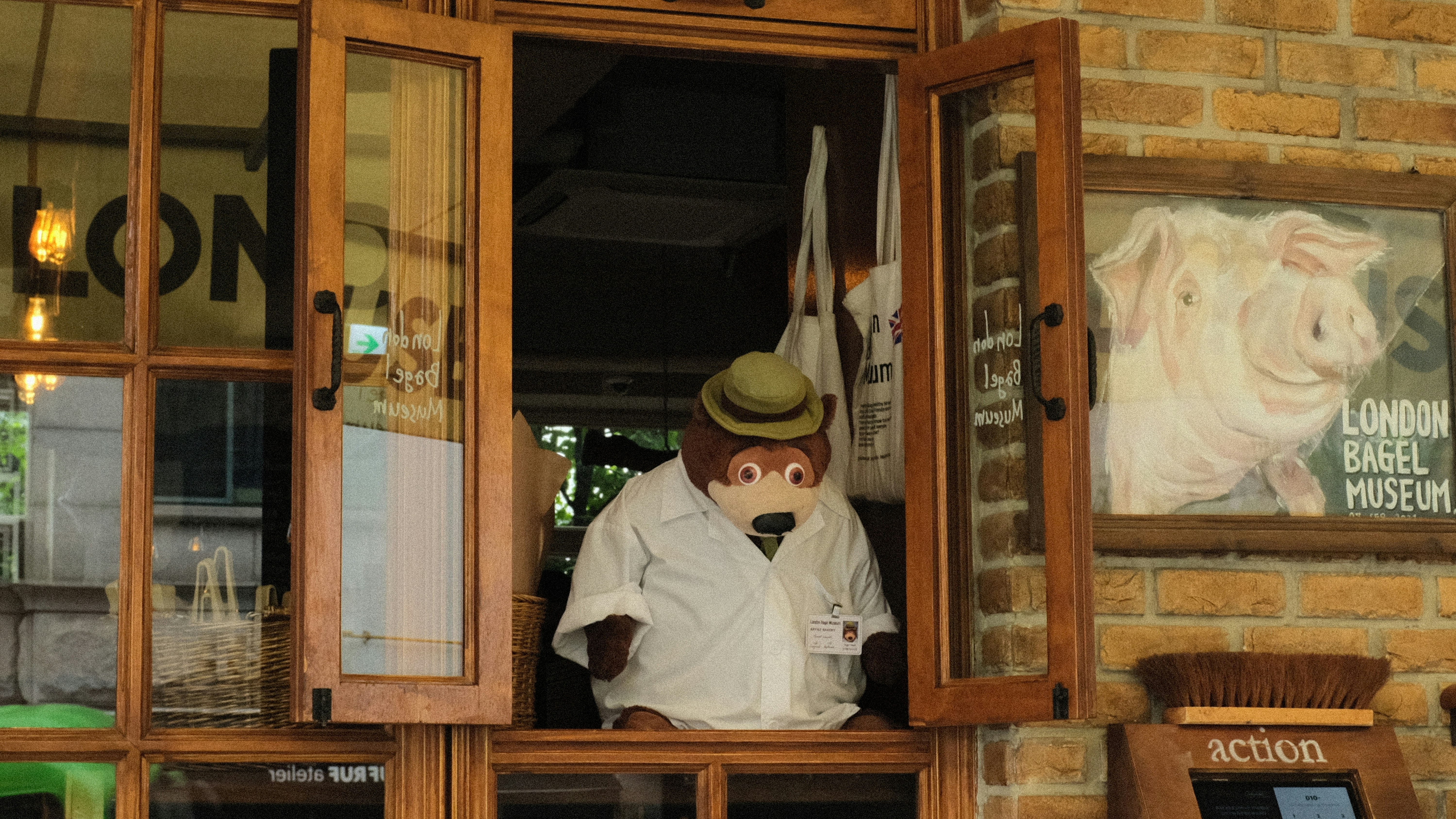 A costumed bear character stands behind an open doorway in a brick-walled indoor setting, framed by glass doors and a poster on the right; the scene resembles a small museum or shop display.
