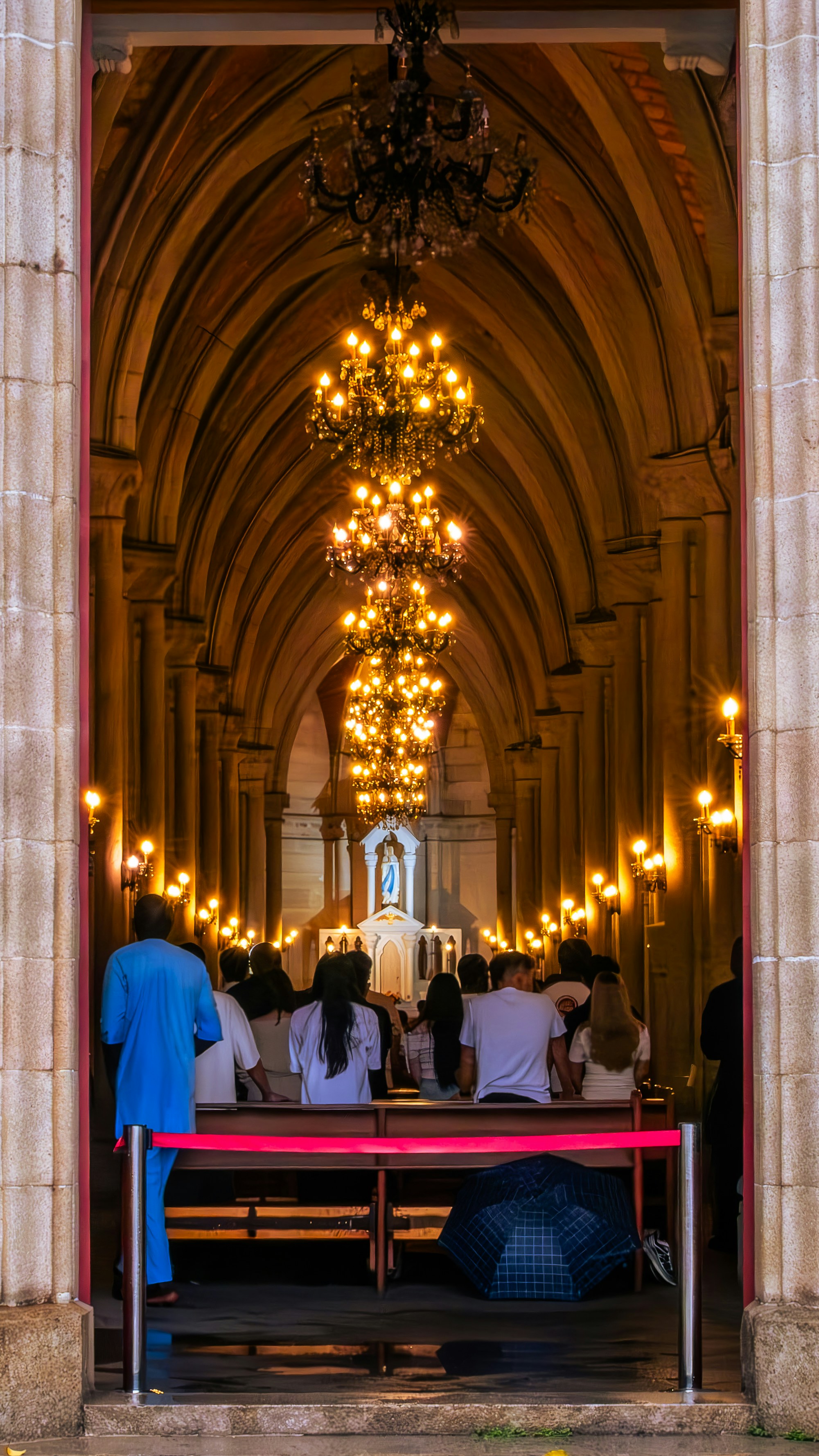 Rows of worshippers fill a grand cathedral aisle, illuminated by ornate chandeliers as light dances along stone arches.