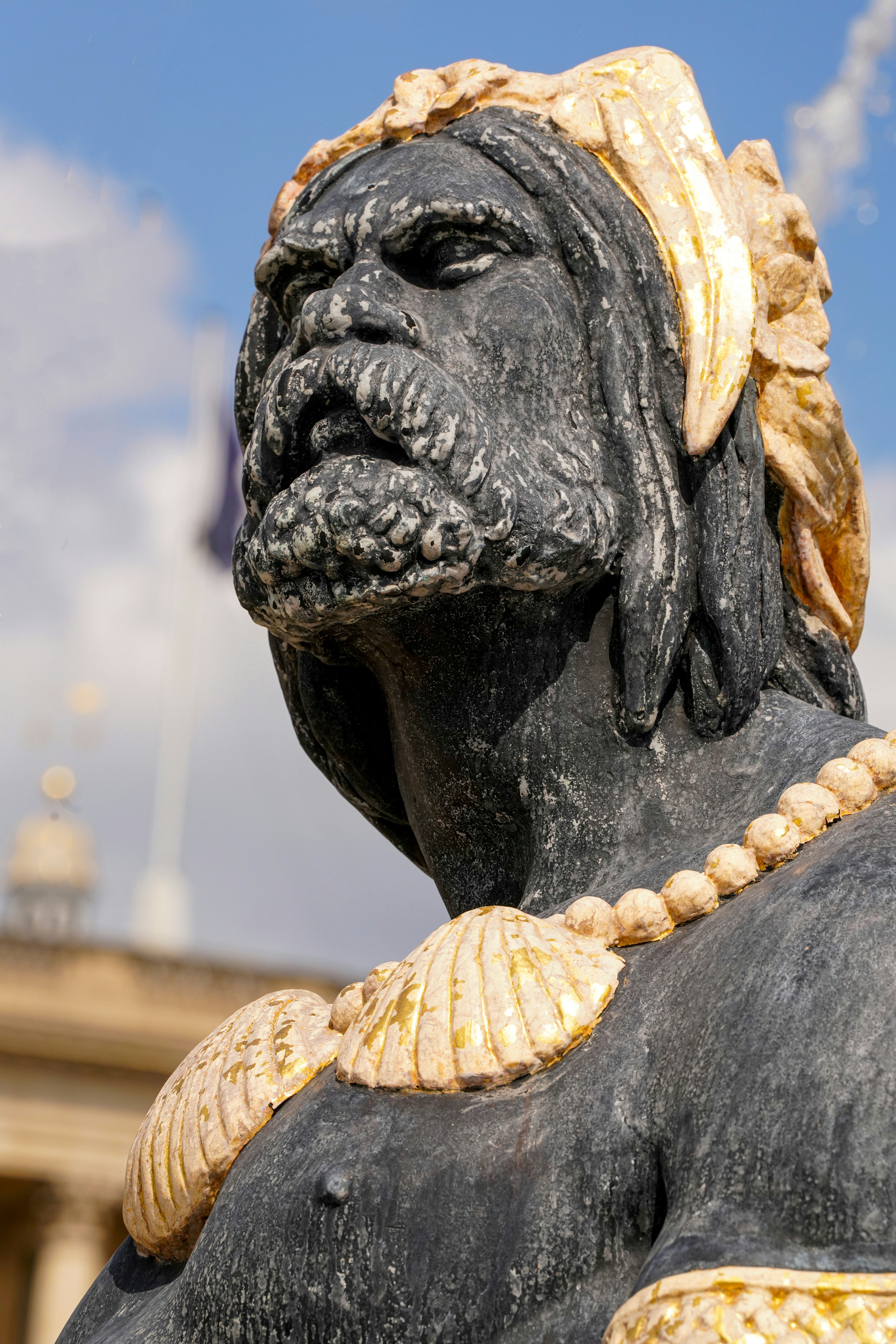 Close-up of a dark stone statue bust with a weathered beard, gold adornments, and a turban; a classical sculpture set against a softly blurred architectural background.