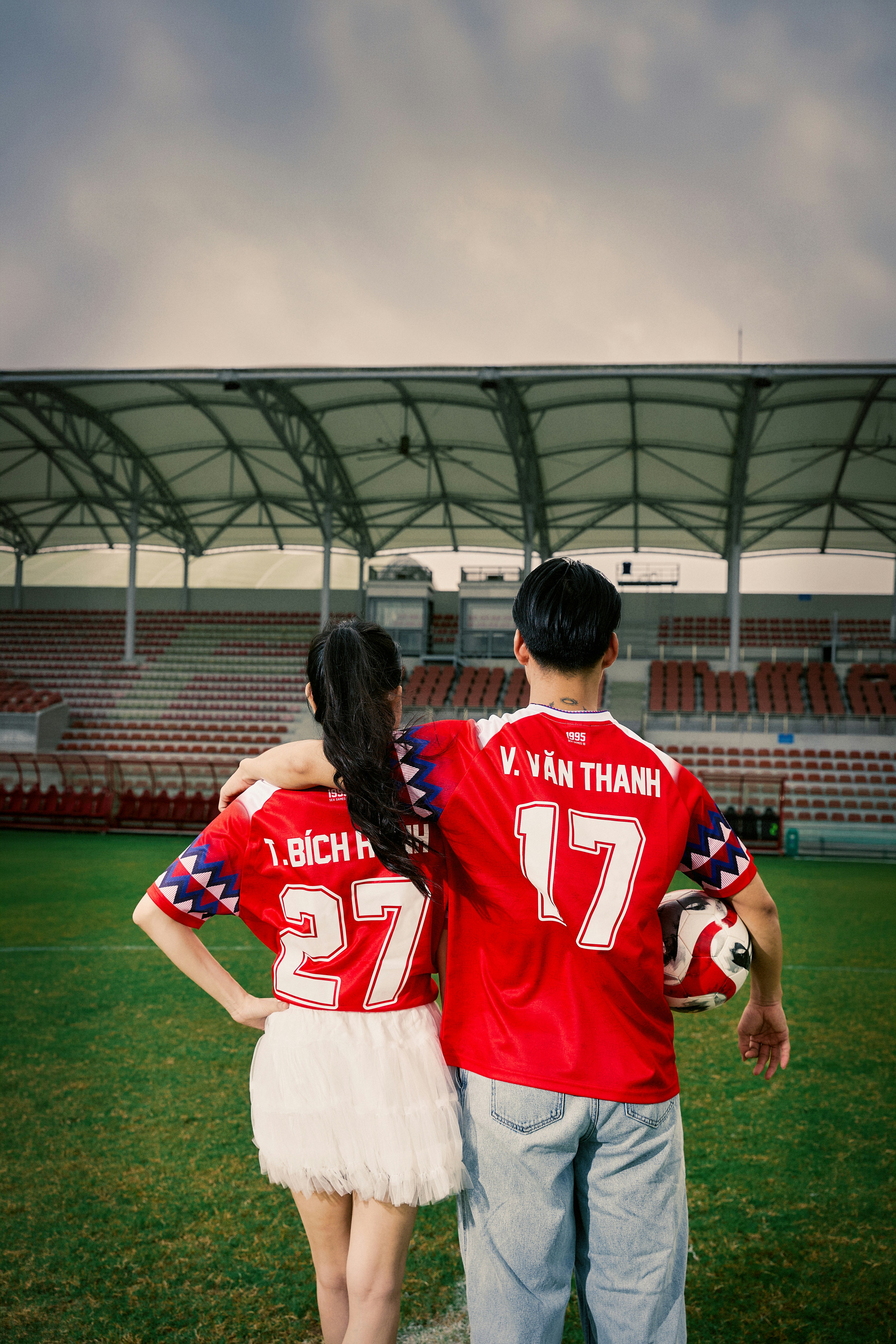 Two teammates with backs to camera in vibrant red jerseys stand under stadium canopy