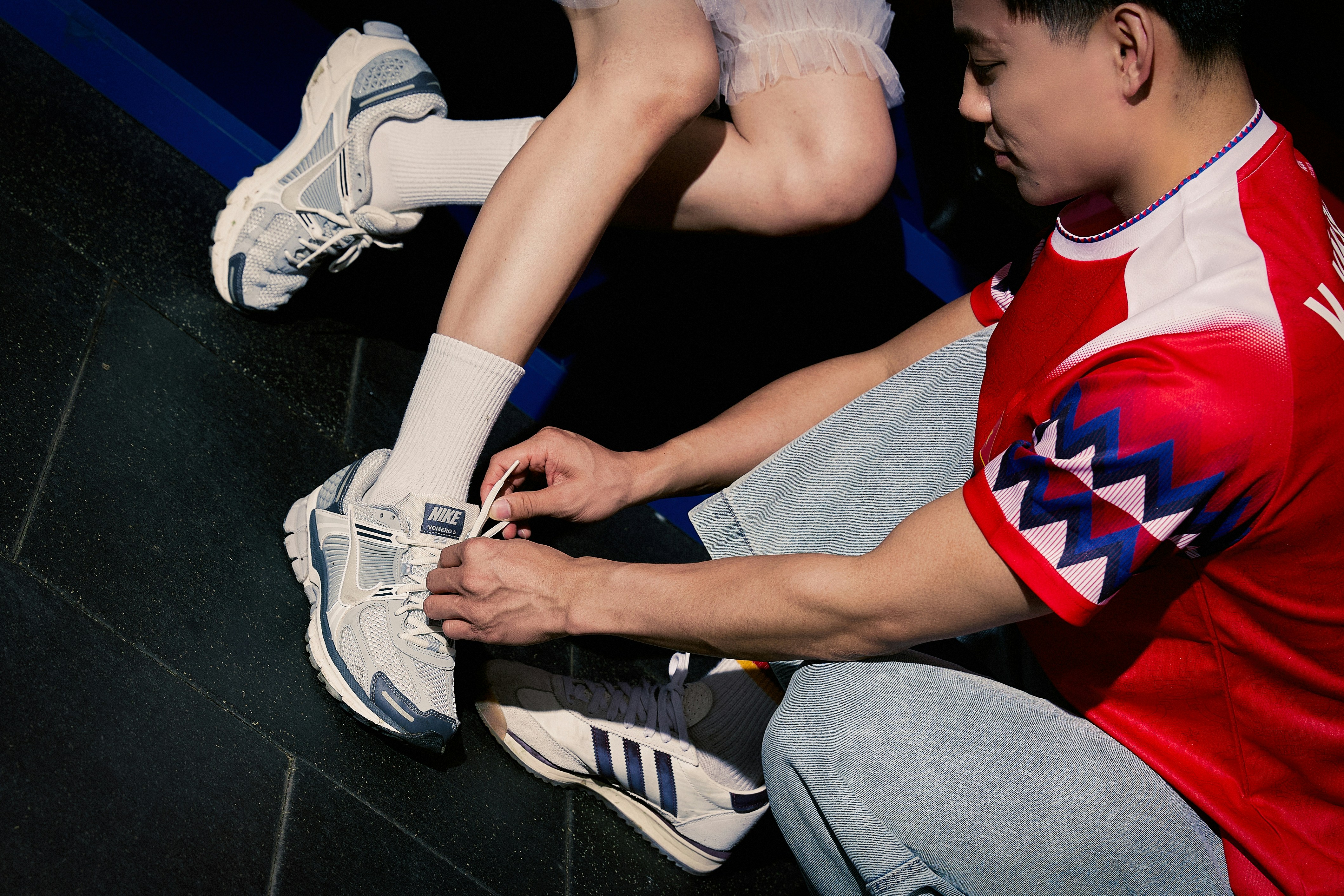 Athlete sits against a dark surface, tying white athletic shoes while another leg crosses in the frame; a red jersey with blue chevrons and gray sweatpants suggest a sports setting.