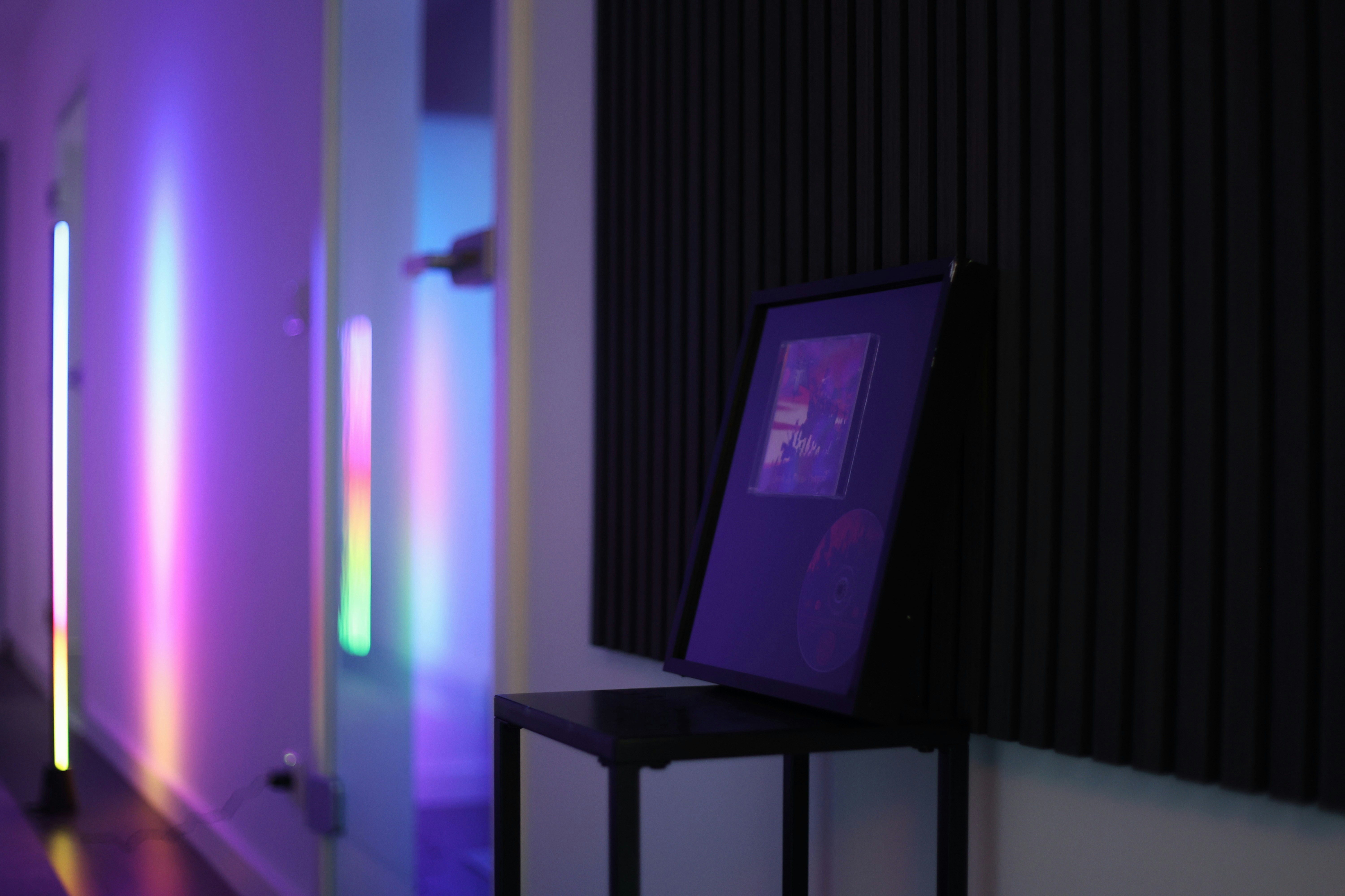 A dim-lit interior scene features a framed display on a small table against a dark, sound-absorbing wall, with neon pastel lights casting a soft glow along a corridor. The setting suggests a studio or gallery space with a moody ambiance.