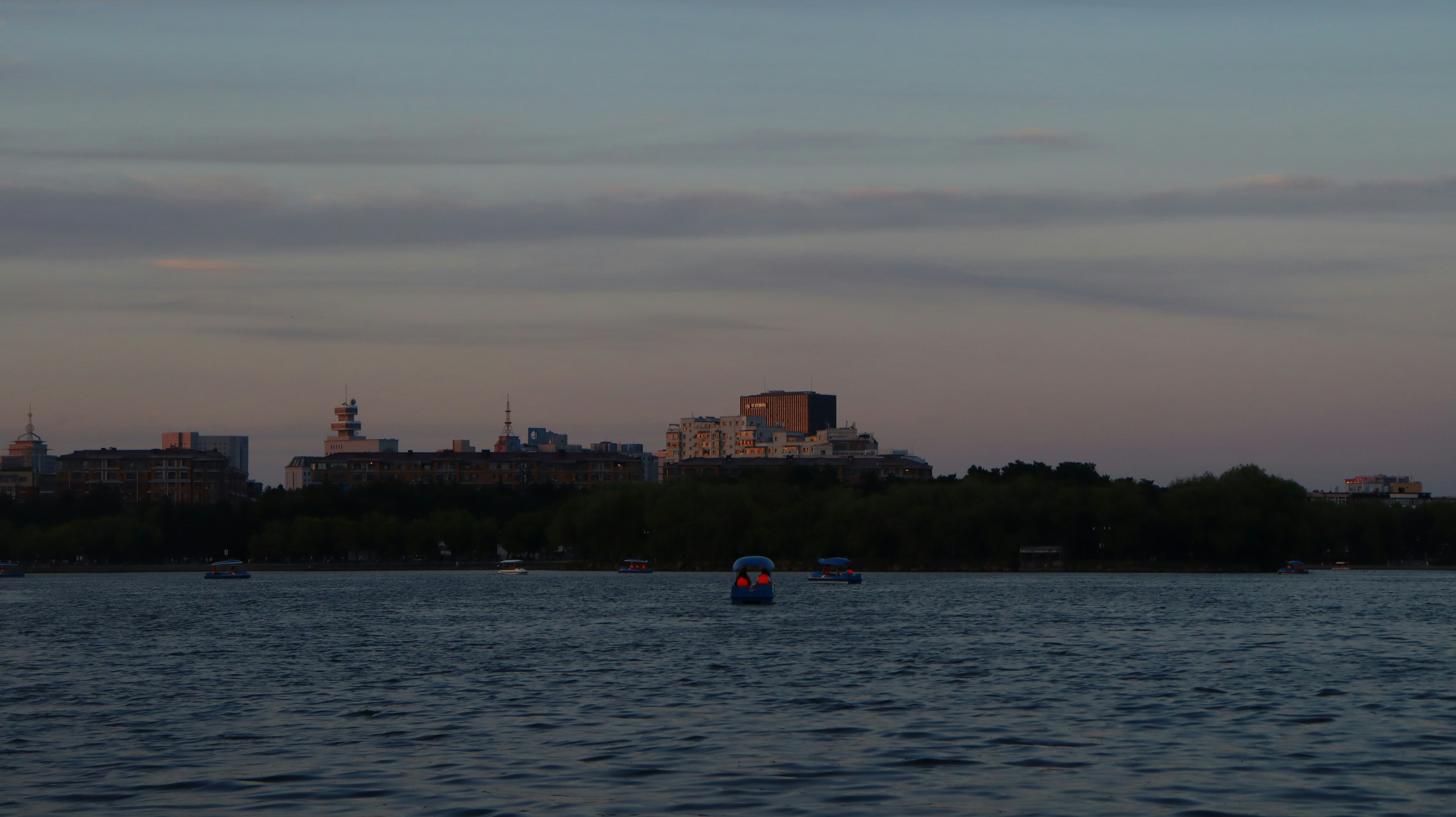 Dusk on a wide river with distant city buildings and small boats dotting the water; a tranquil urban waterfront at twilight.