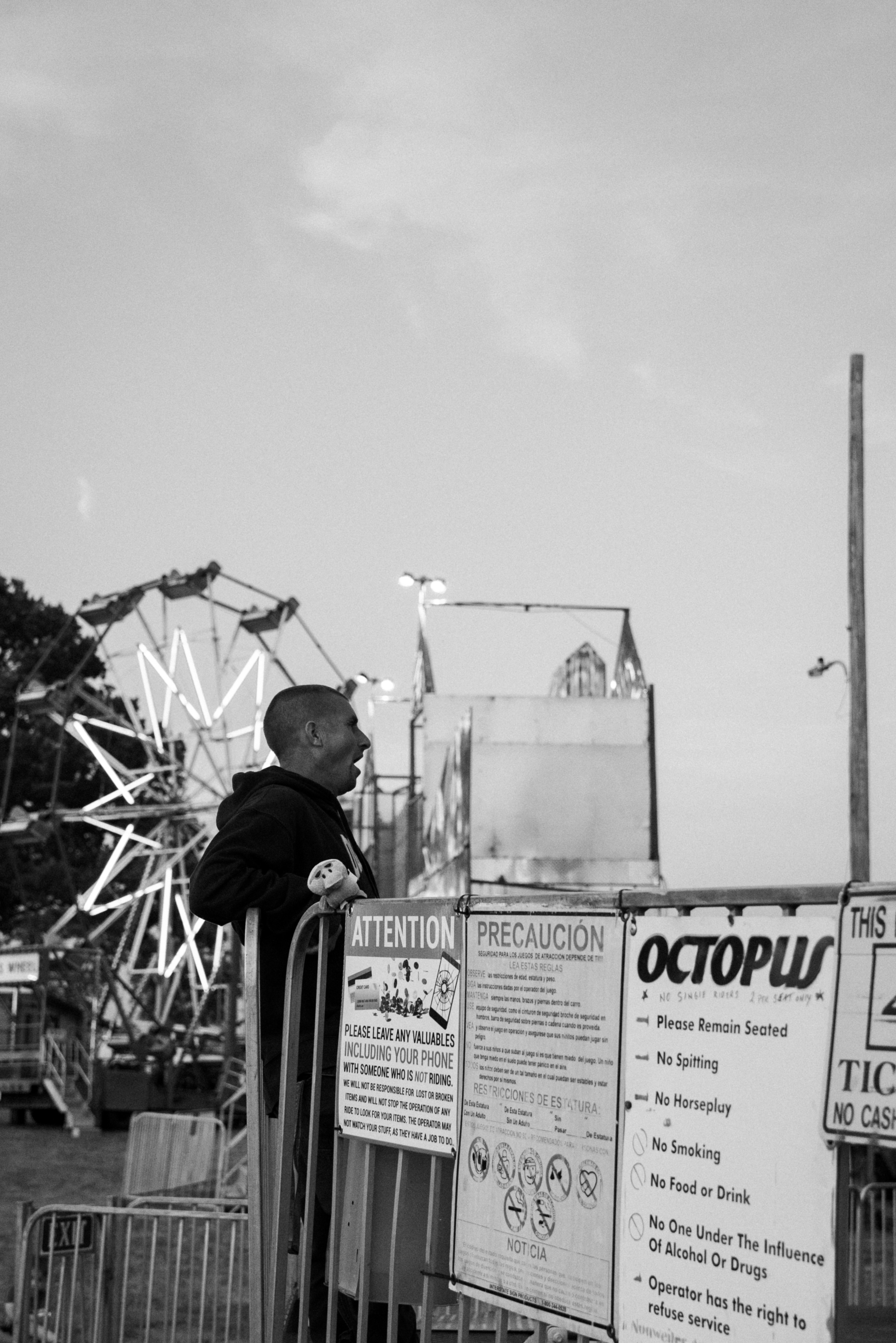 Man stands behind a barricade at a fairground entrance, signage and a rotating ride structure in the background; monochrome keeps focus on the boundary between the public space and the attraction area.