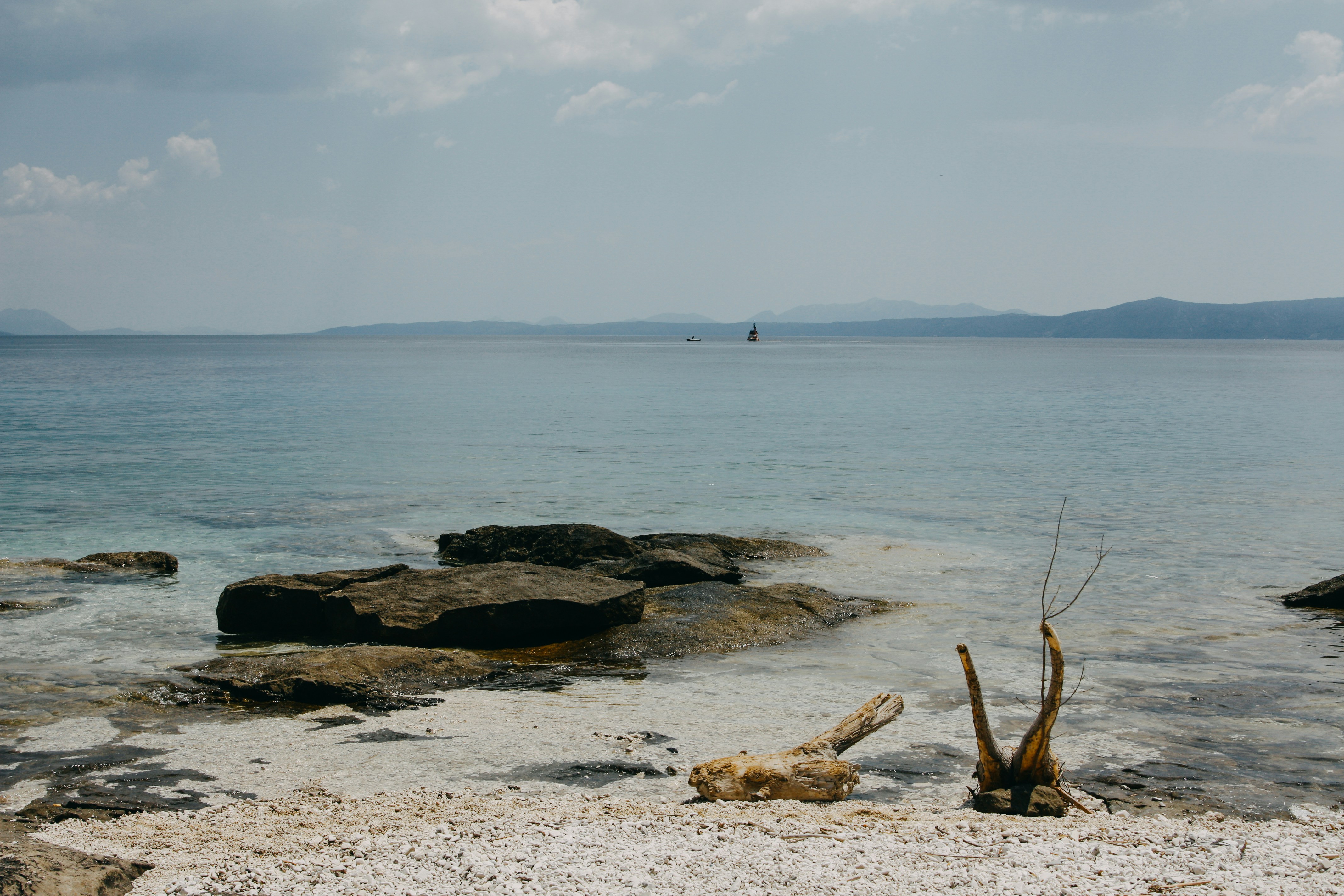 Sunlit pebbled beach with scattered driftwood and dark rocks along the calm sea; distant horizon and boats visible beyond the shoreline.