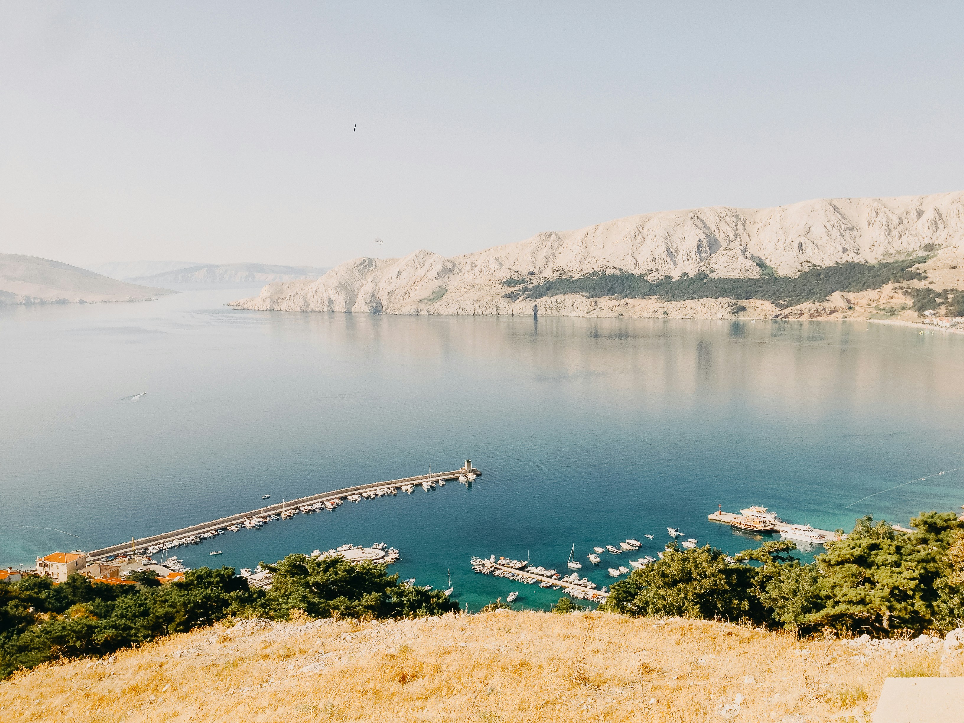 Calm harbor with a wooden pier extending into turquoise water, framed by rocky hills and a golden grassy foreground.