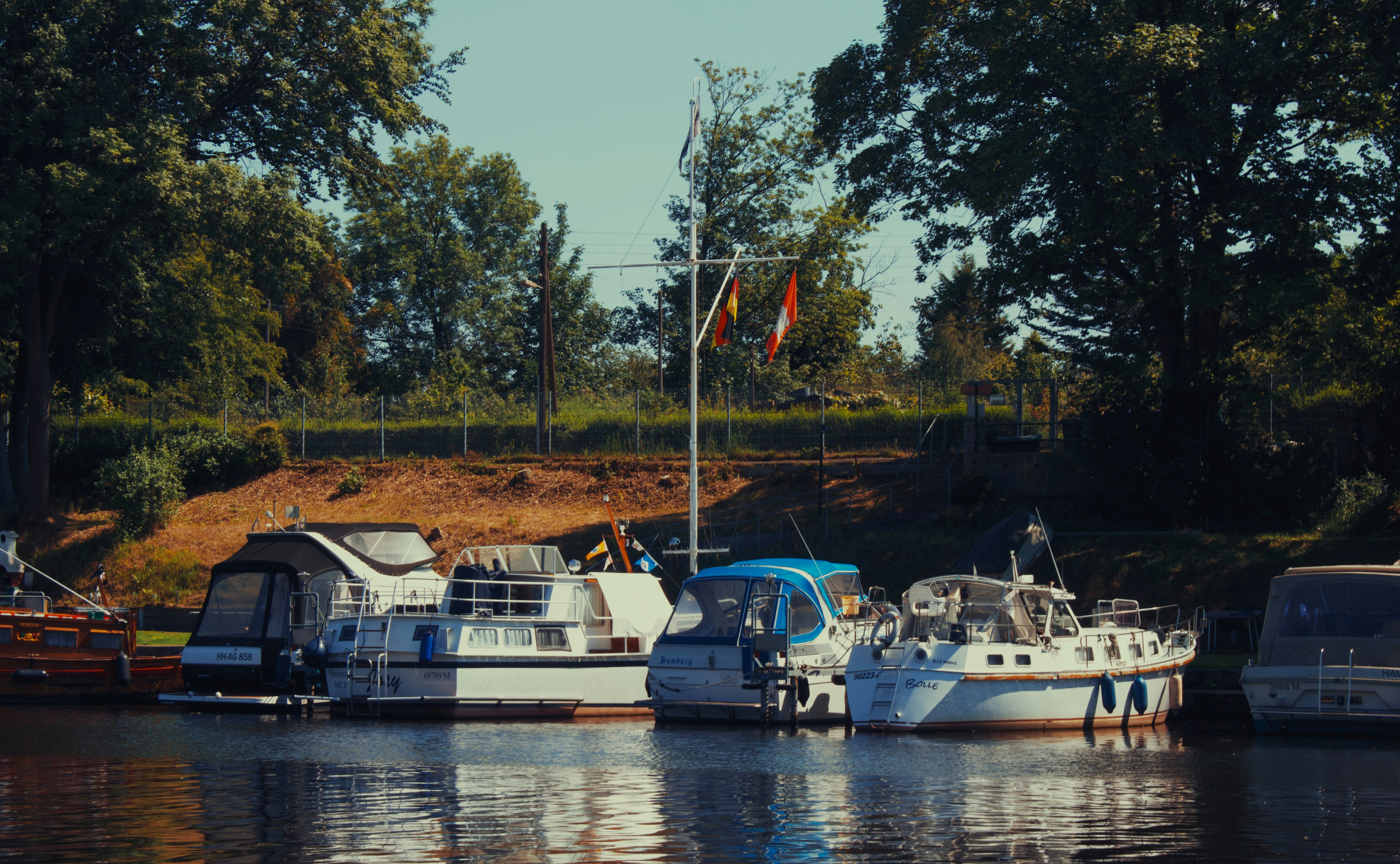  Boats docked along a calm river with trees and flags in the background; a tranquil harbor scene with shaded water reflections. 