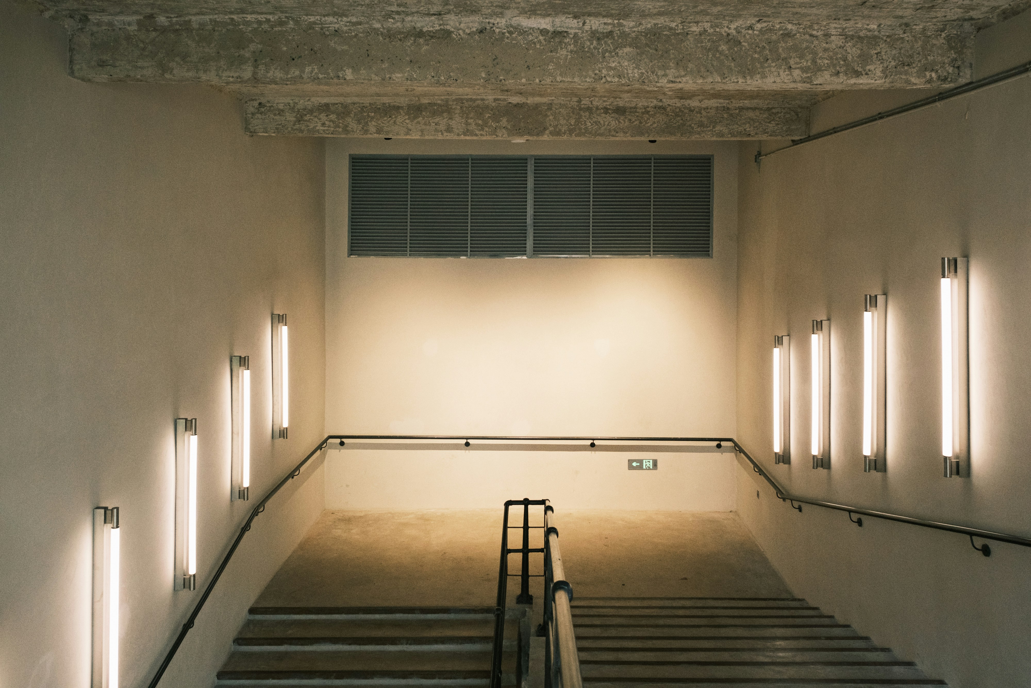 Minimalist stairwell with warm vertical lighting along walls, leading toward a dim exit; architectural concrete and metal handrails create a somber, atmospheric space.