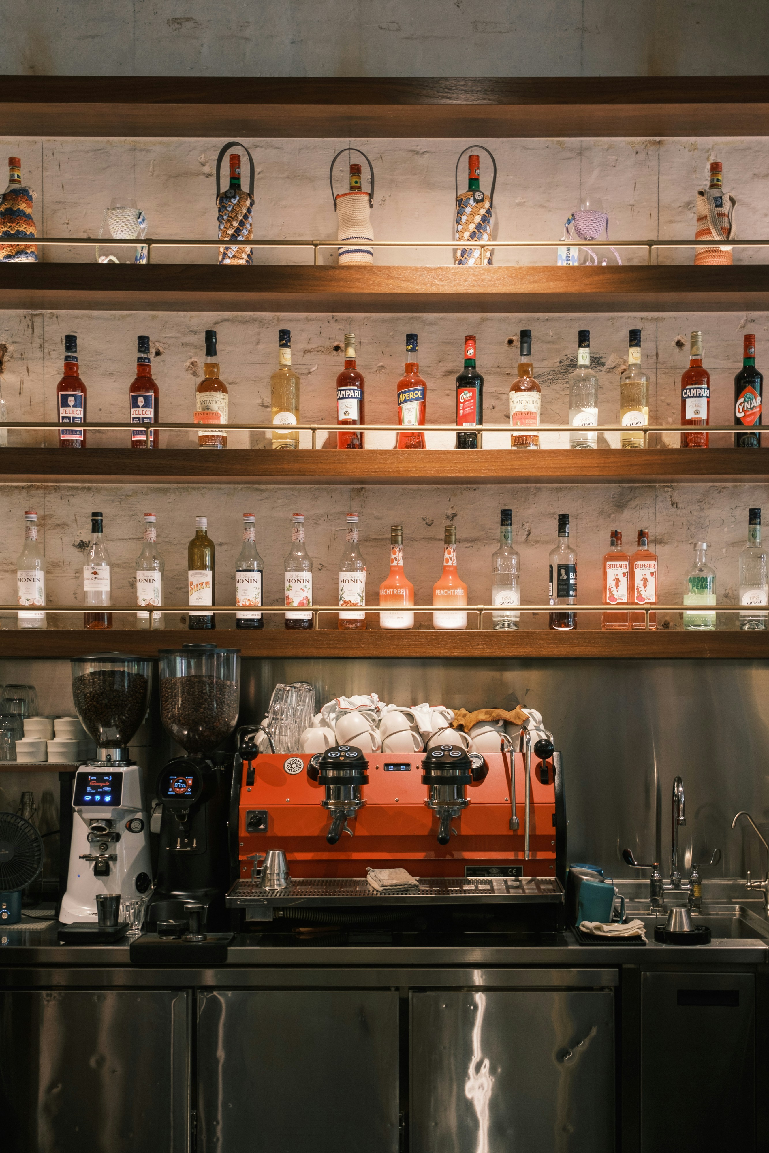 Barista counter with an orange espresso machine, bottles on wooden shelves, and coffee grinders in the foreground. A warm-toned, cafe-style display with metal surfaces and soft lighting.