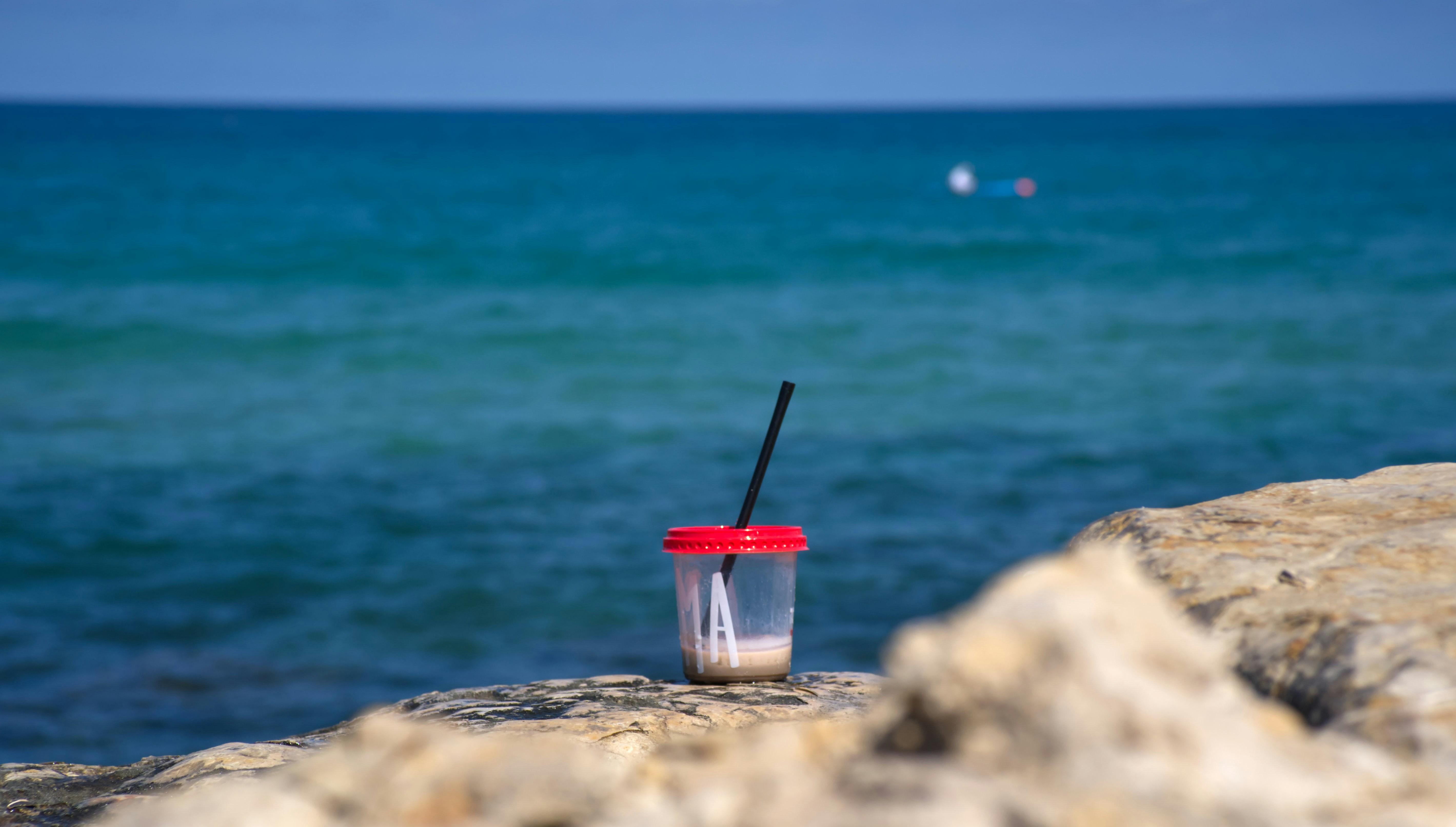 A small cup with a red lid sits on sunlit rock along a turquoise sea shoreline.