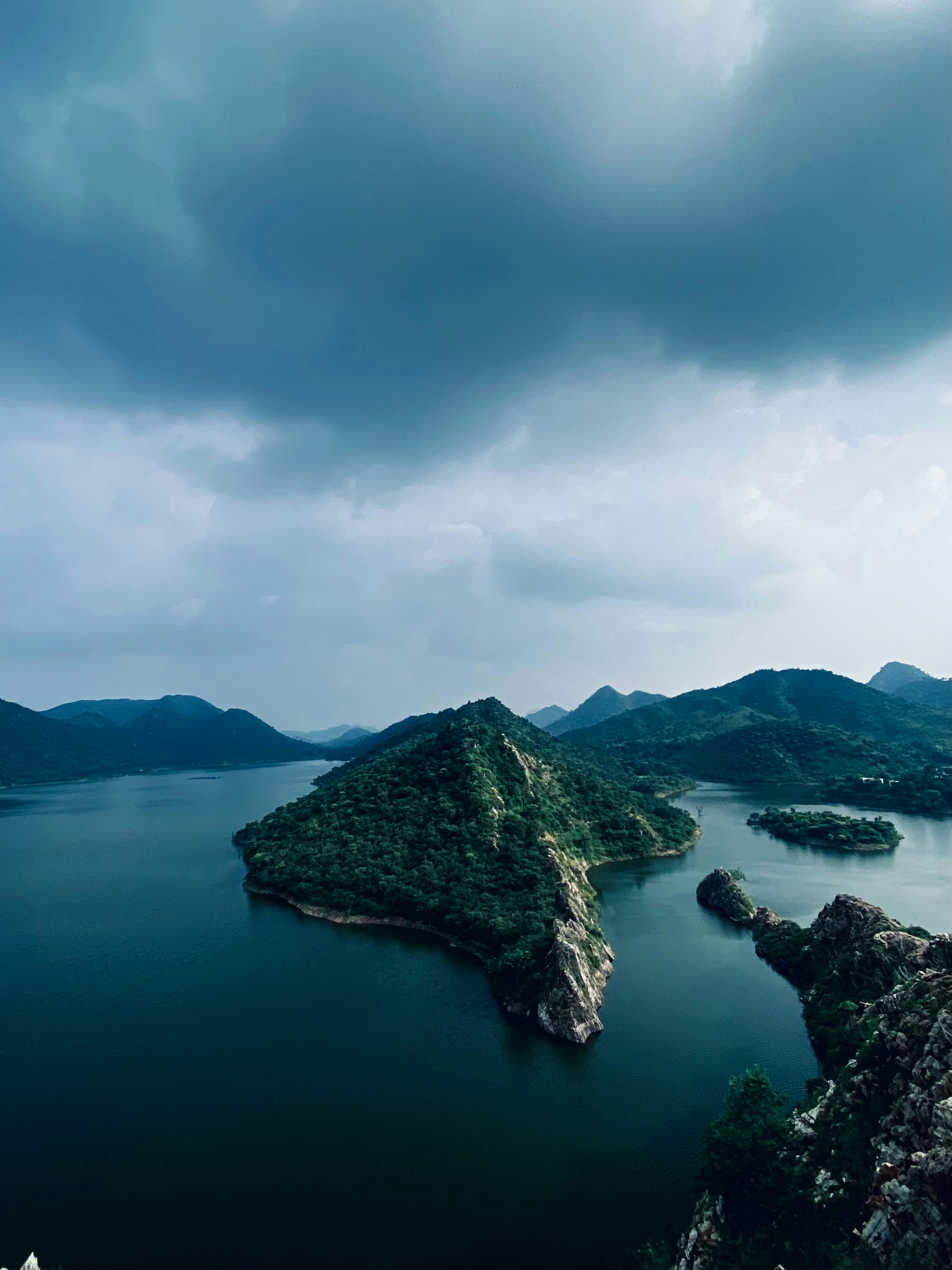 Verdant island in a calm bay under a dramatic, cloud-filled sky, with surrounding rugged shoreline and distant hills.