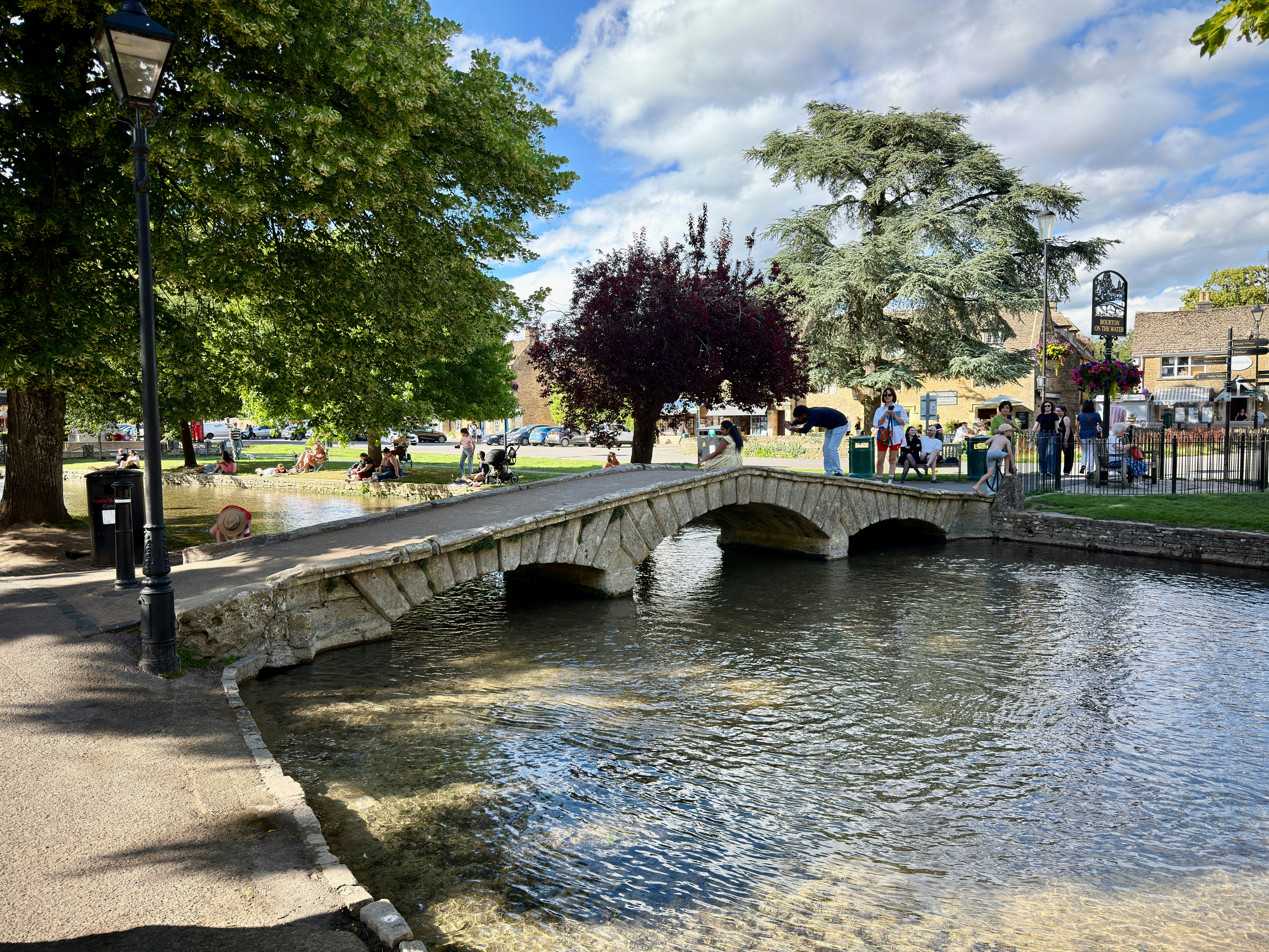 Shallow canal with a stone arch bridge and nearby parkgoers in a sunlit town square, trees lining the water and a bright sky.