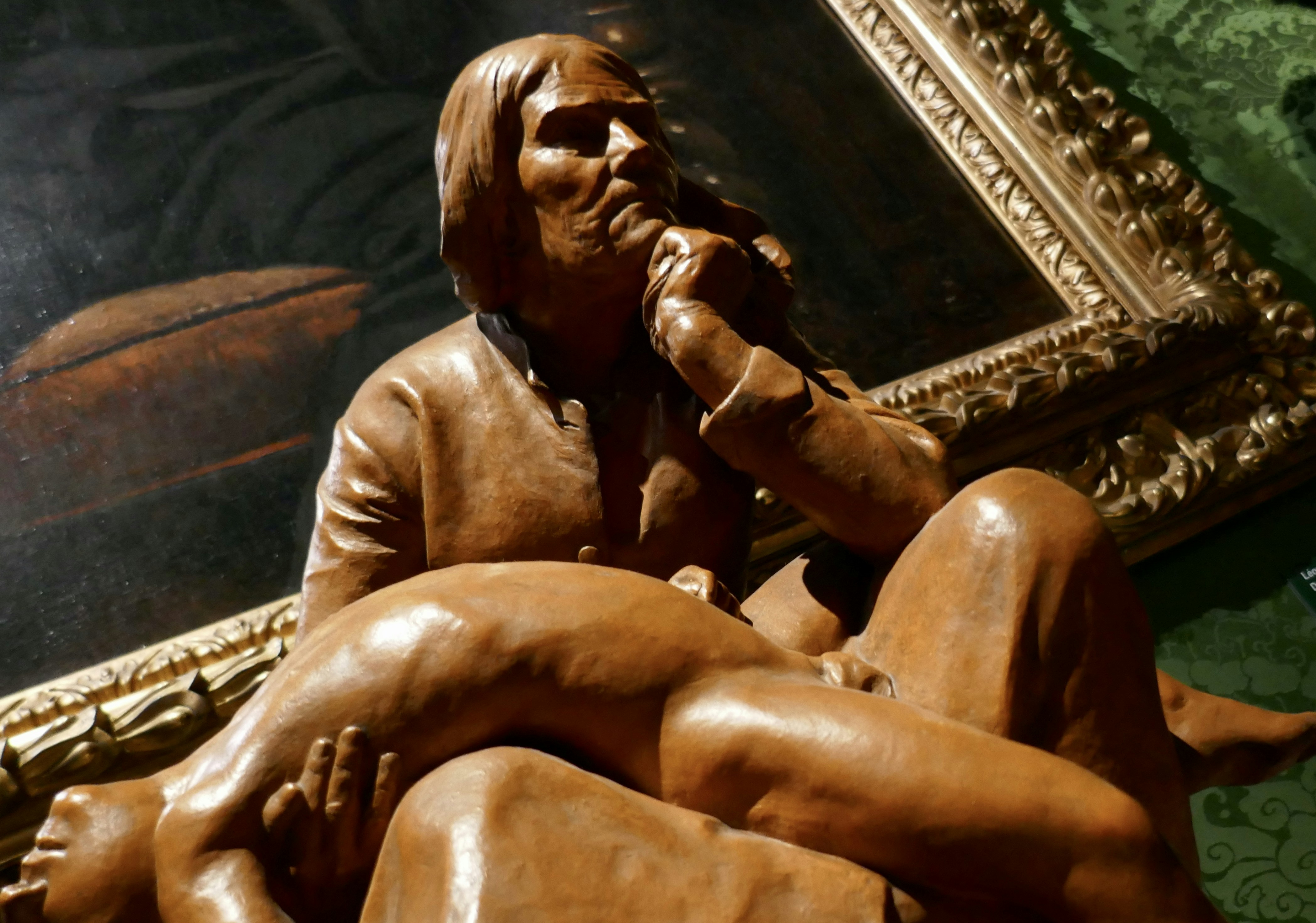 Sculpted bronze statue captured against an ornate frame and green backdrop, highlighting its reflective surface and contours. The composition centers the figure amid the decorative surroundings.