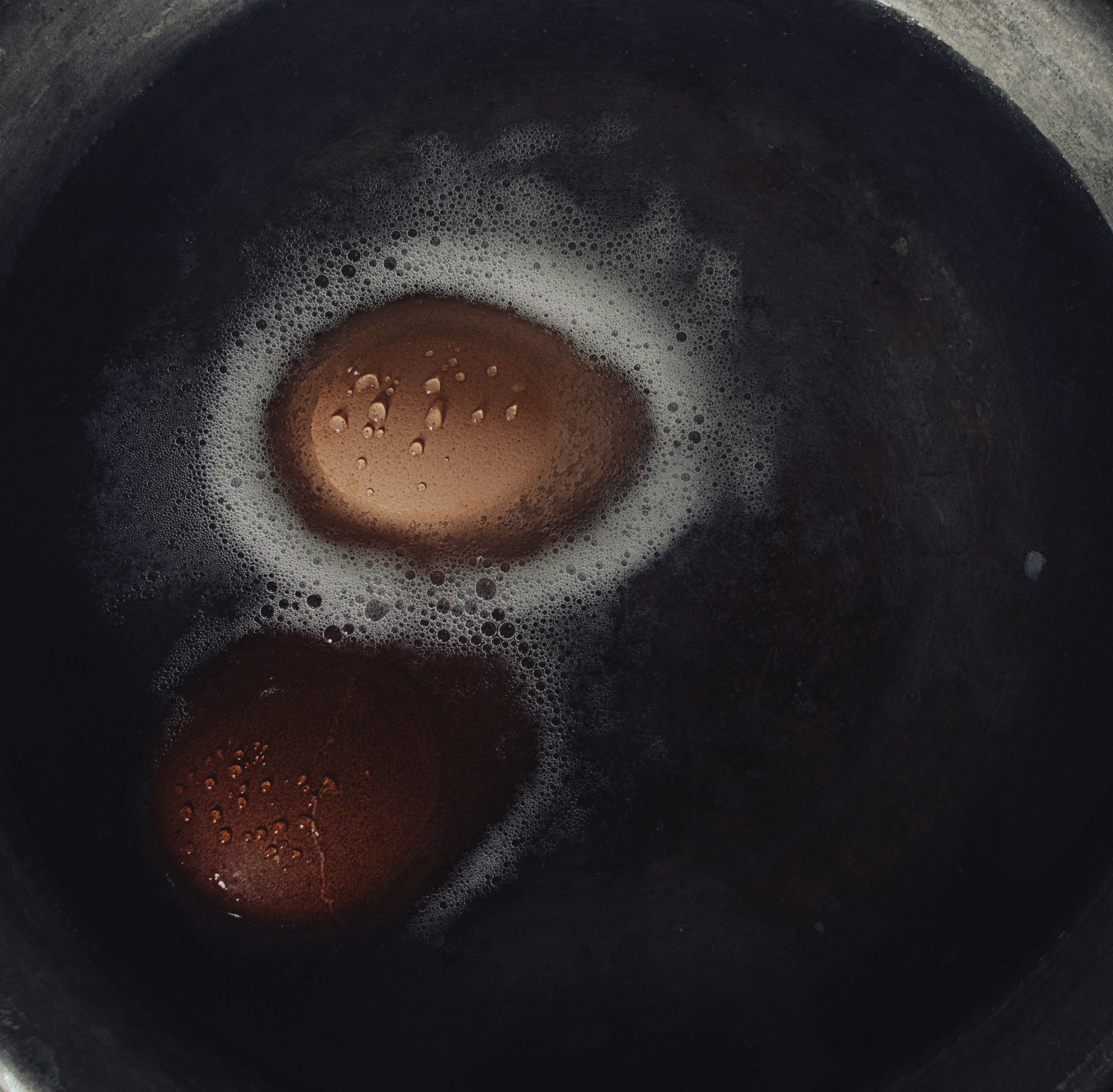 Two eggs simmer in a dark pot with frothy bubbles forming around them, suggesting a quiet cooking moment.