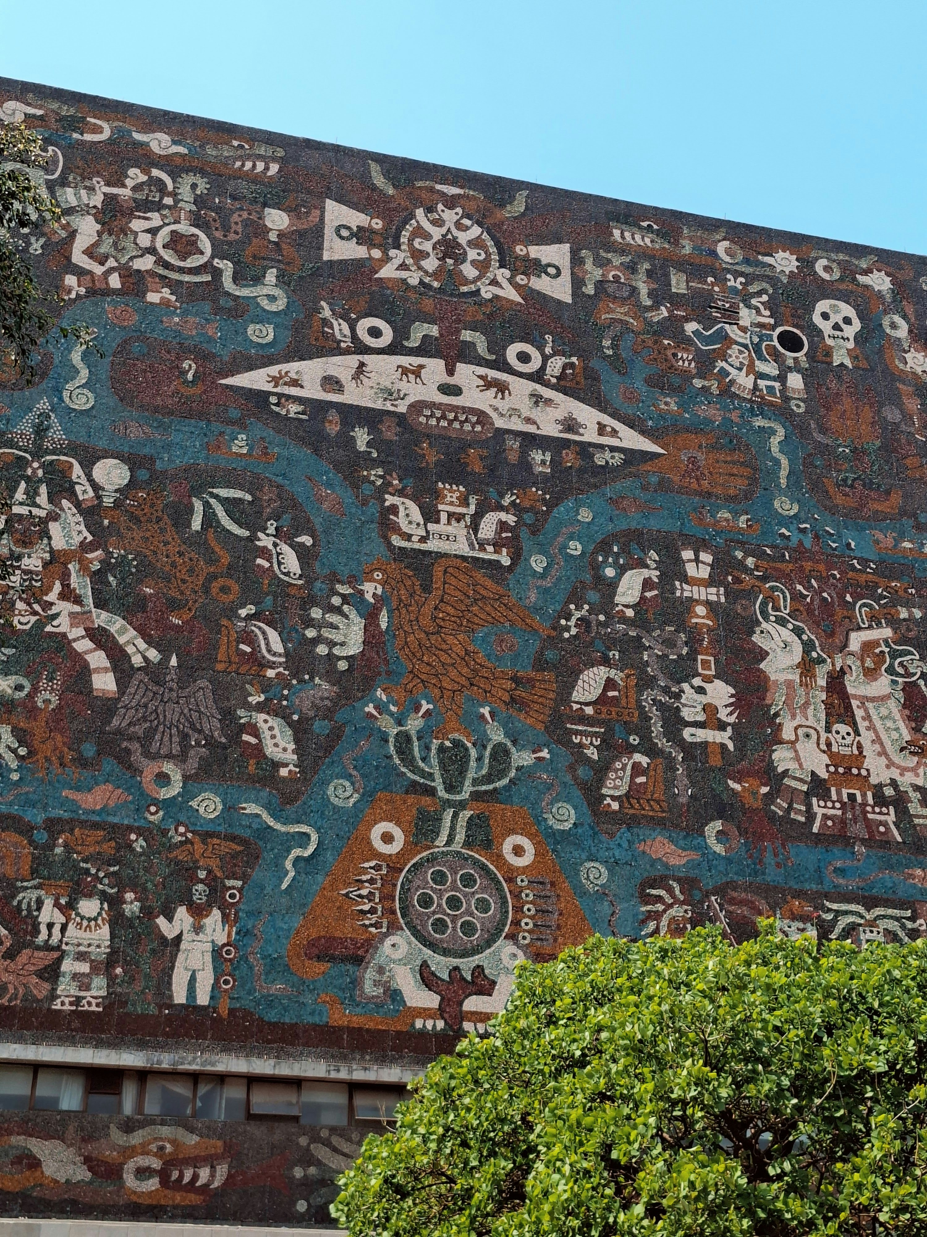Detailed mosaic mural on a building with green foliage.
