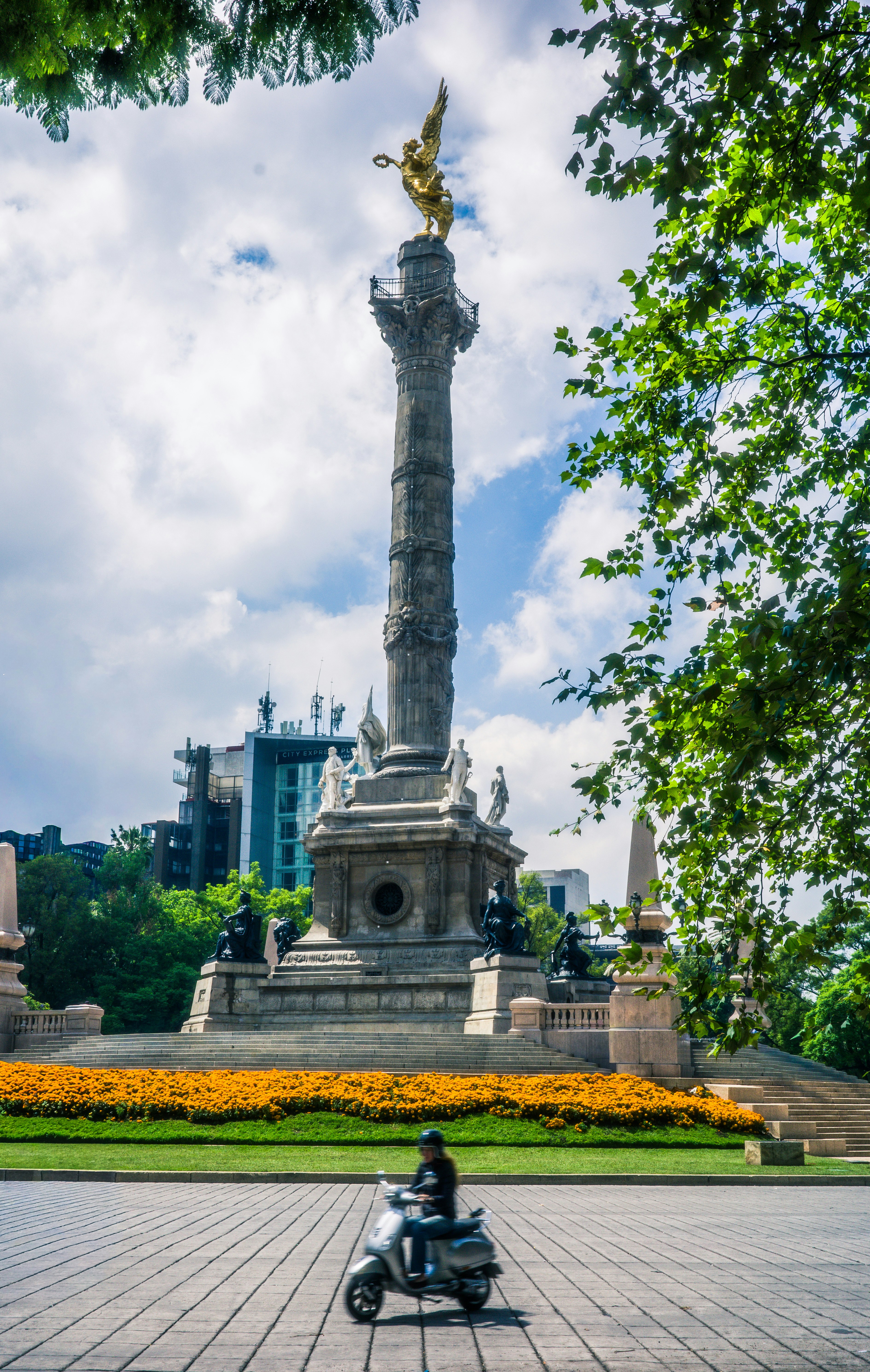 Sculpted monument rising above a plaza with a scooter rider passing in the foreground; a bright blue sky with clouds behind and manicured flower beds at the base.