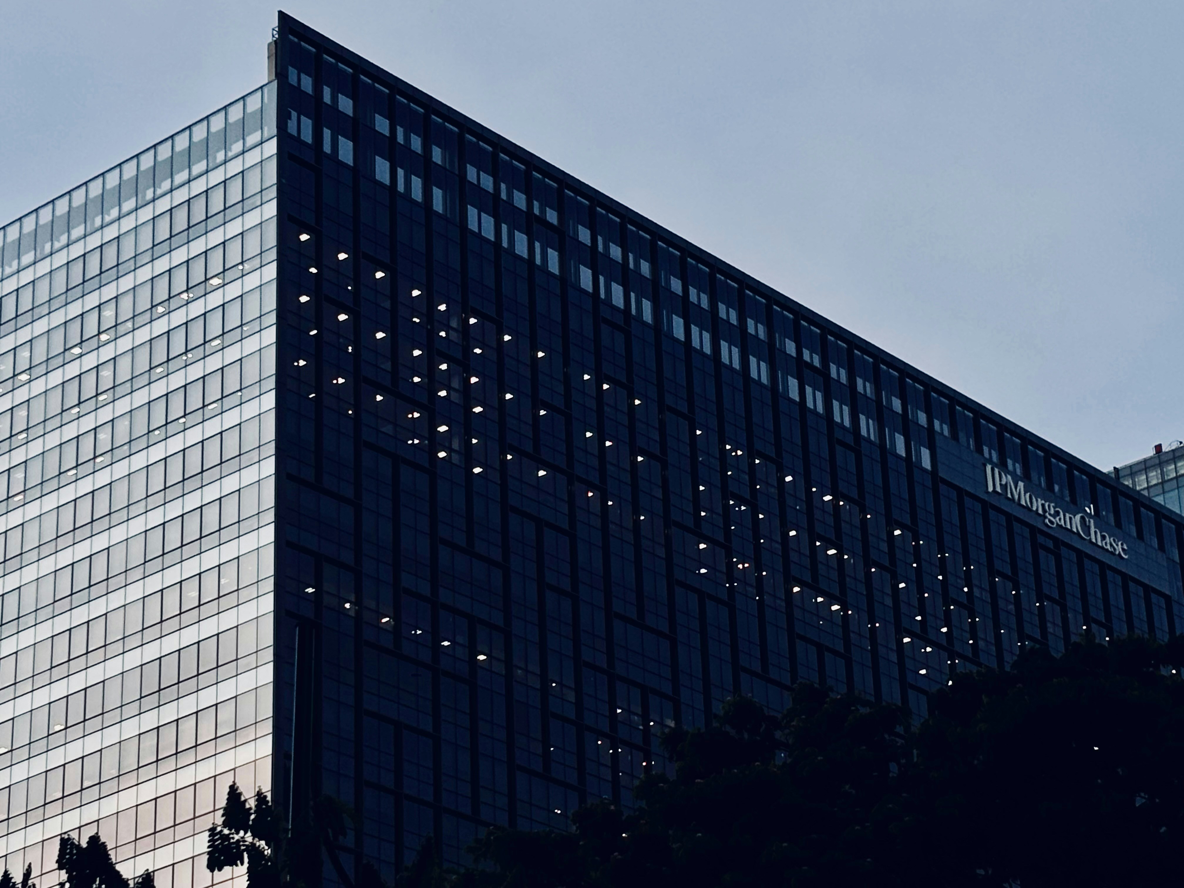Glass-clad office building with illuminated interior lights at dusk; dark trees in foreground and a pale sky, highlighting modern architecture.