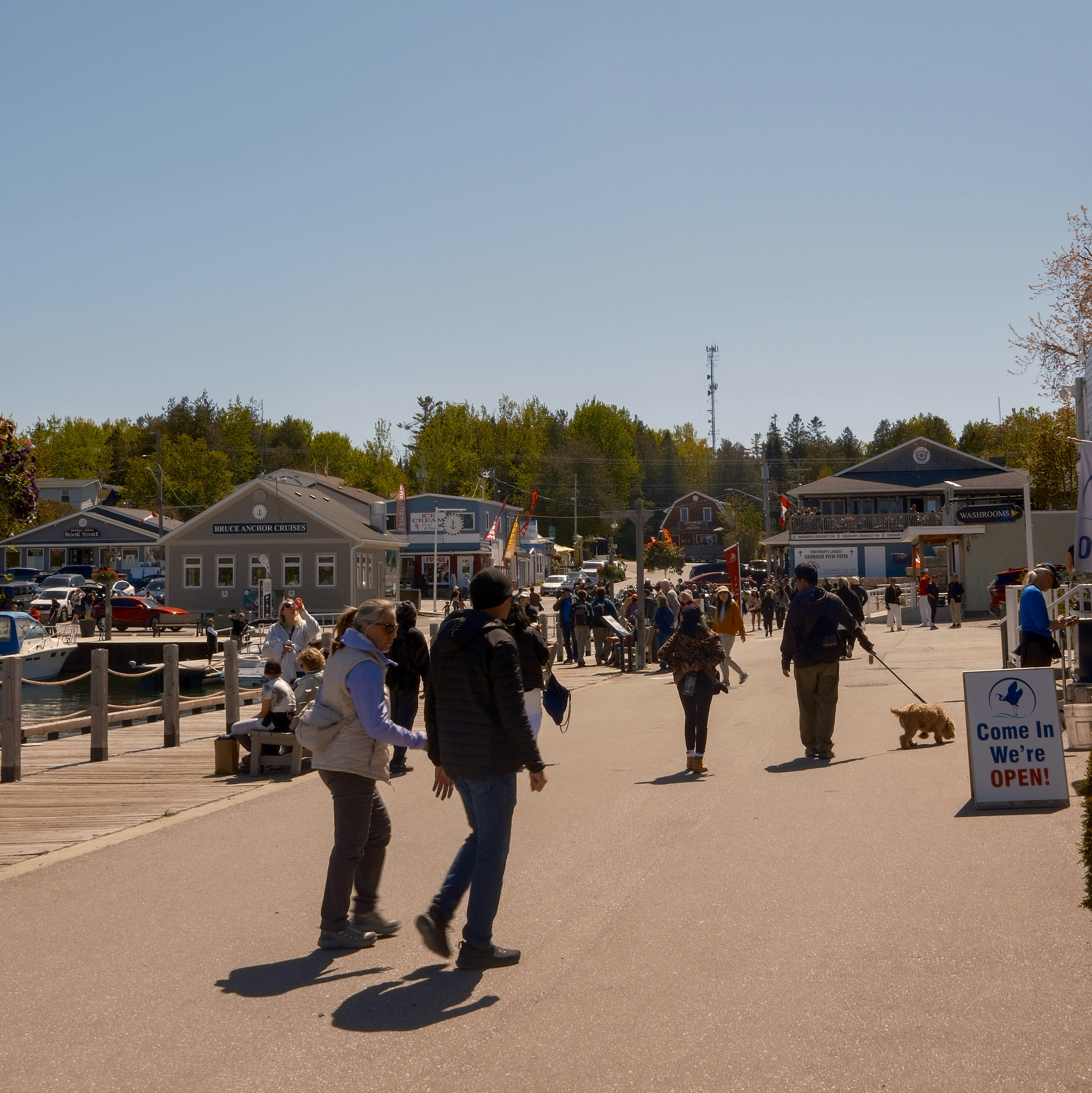 A bright, high-energy street scene capturing the lively tourism culture of a coastal resort village. The composition tracks visitors exploring a harbor road lined with excursion booking offices and boardwalk shops under a crisp sky. A bold, graphic welcome sign on the right grounds the image, giving it a strong commercial, editorial feel. The native contrast and vibrant daylight tones are rendered flawlessly by the sensor. This lively shot is a perfect fit for travel networks, local business sponsors, and commercial tourism campaigns.