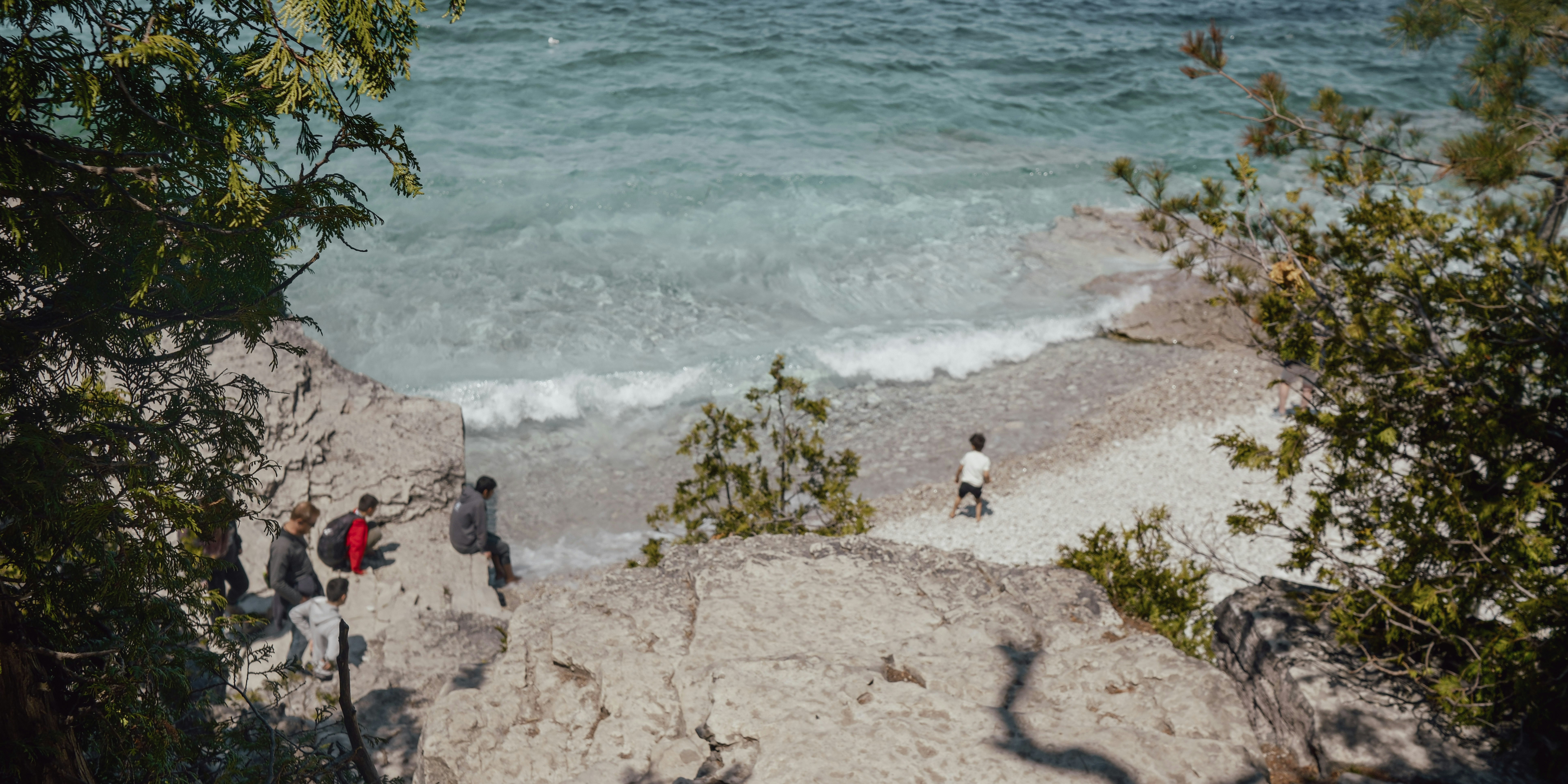 A high-angle documentary frame looking down from a rocky cliff onto a rugged pebble beach. Small groups of travelers sit along the weathered stones, while a lone figure explores the shoreline as gentle white waves wash ashore. The vast expanse of clear, pale blue water stretches out into the upper half of the composition. This unedited slice of outdoor culture serves as a versatile lifestyle asset for activewear campaigns, travel channels, and publications tracking human experiences in the wild.
