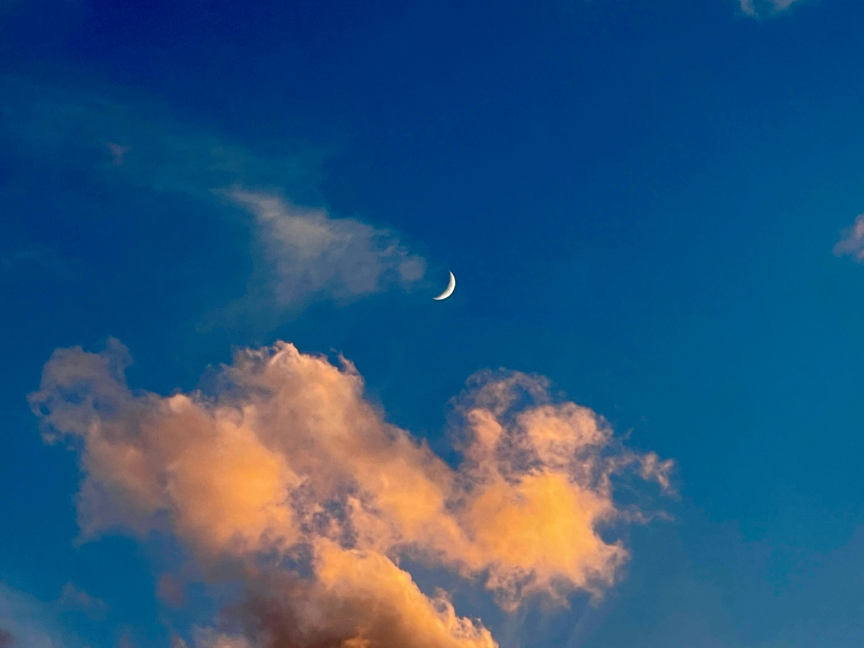 Crescent moon above warm clouds in a calm blue sky; minimal composition with soft, vivid sunset hues and wispy clouds.