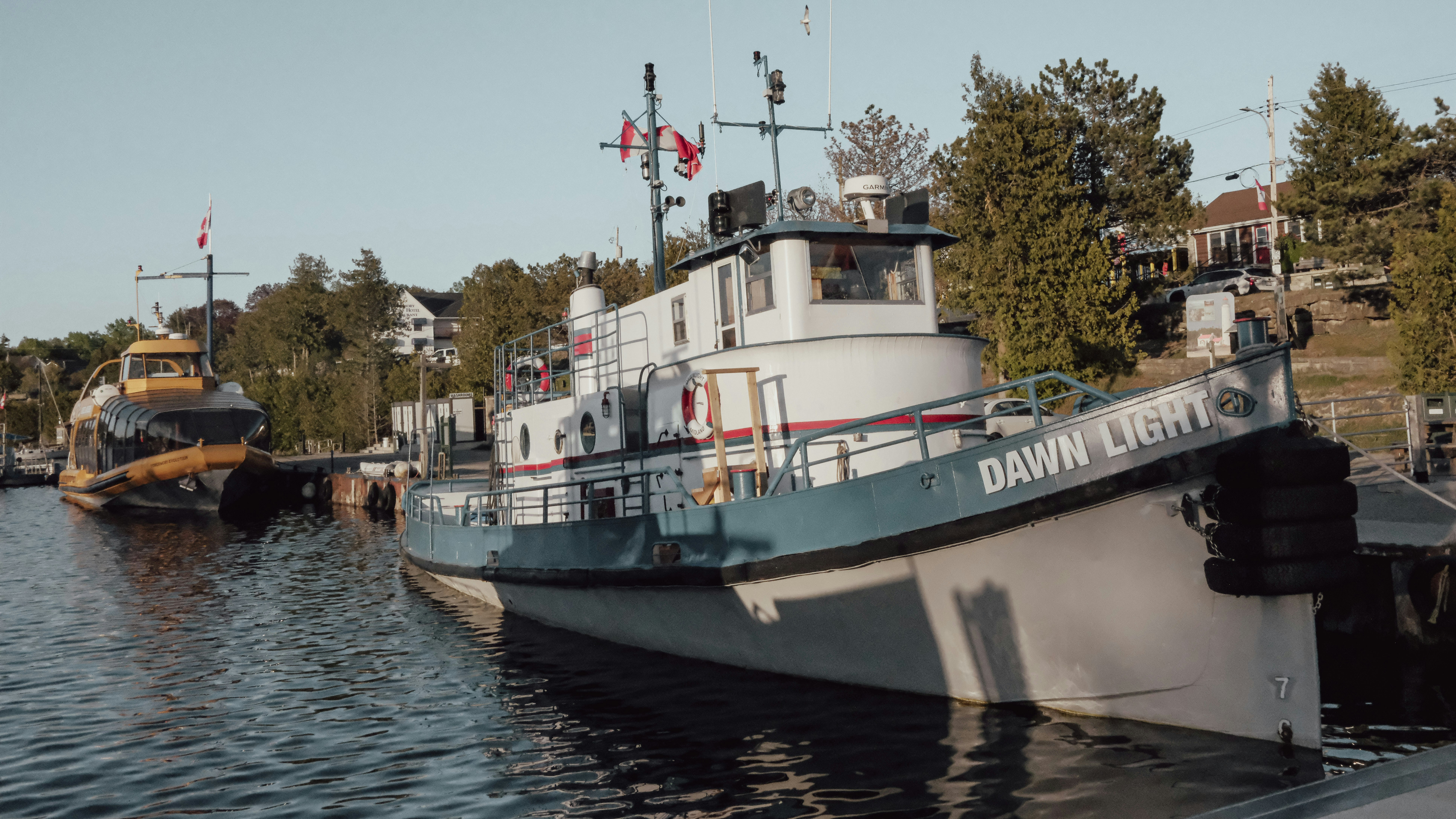 A wide shot showing two different kinds of boats in the harbor. In the front, there is an old white fishing boat tied to the pier. Farther back in the water, you can see a bright yellow tour boat for tourists. I love how the camera captures the old heritage next to the modern travel lifestyle. The colors look very warm and natural without any computer editing. This is a great storytelling image for regional tourism boards or travel channels.