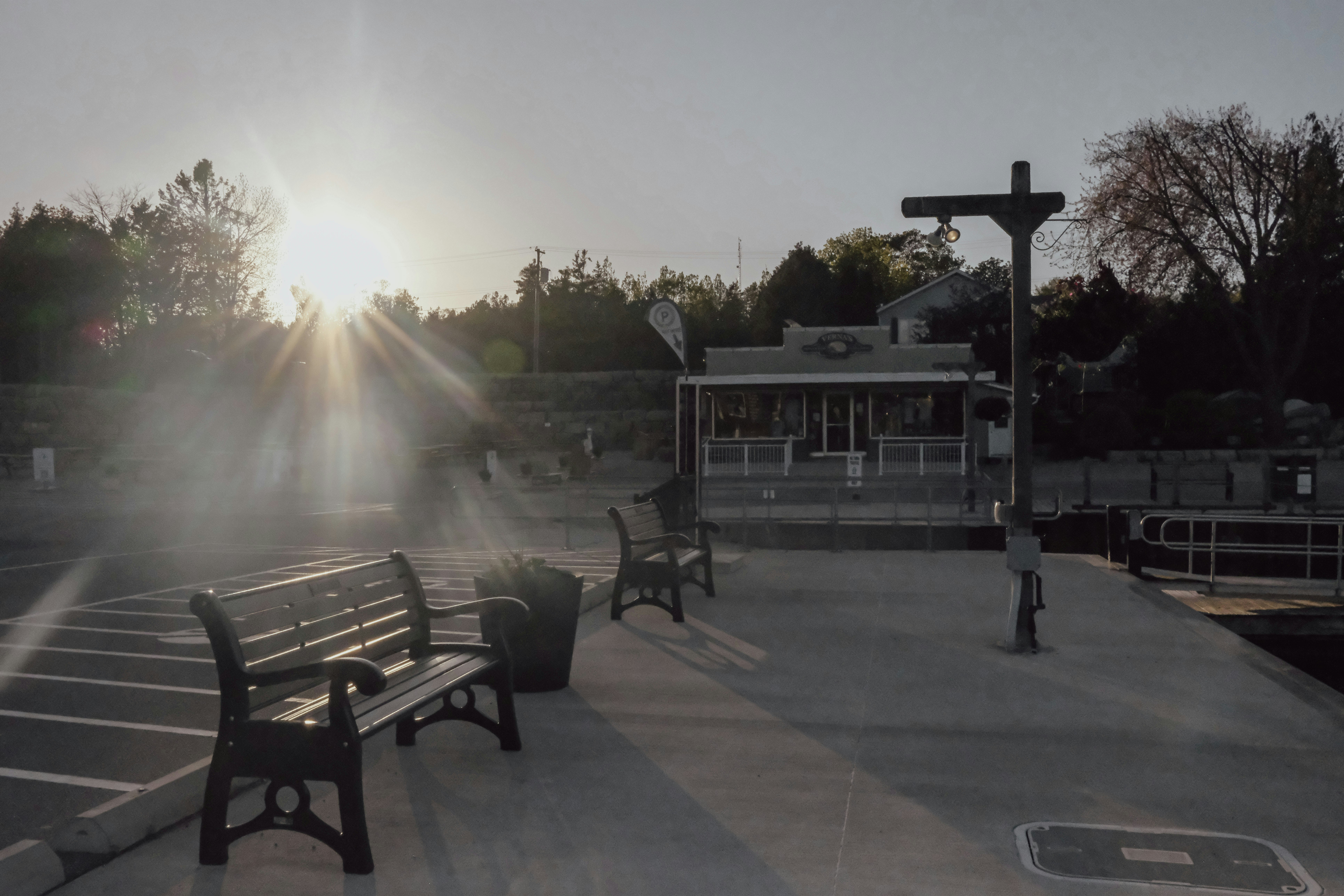 A beautiful morning view of a quiet town square right at sunrise. The sun is shining very bright through the trees, creating a nice lens flare and long shadows on the concrete path. There are empty wooden benches facing a small local shop in the back. The camera handled the bright sunlight perfectly without losing the dark details. It has a great cozy feel for hospitality brands, travel blogs, or lifestyle sponsors.