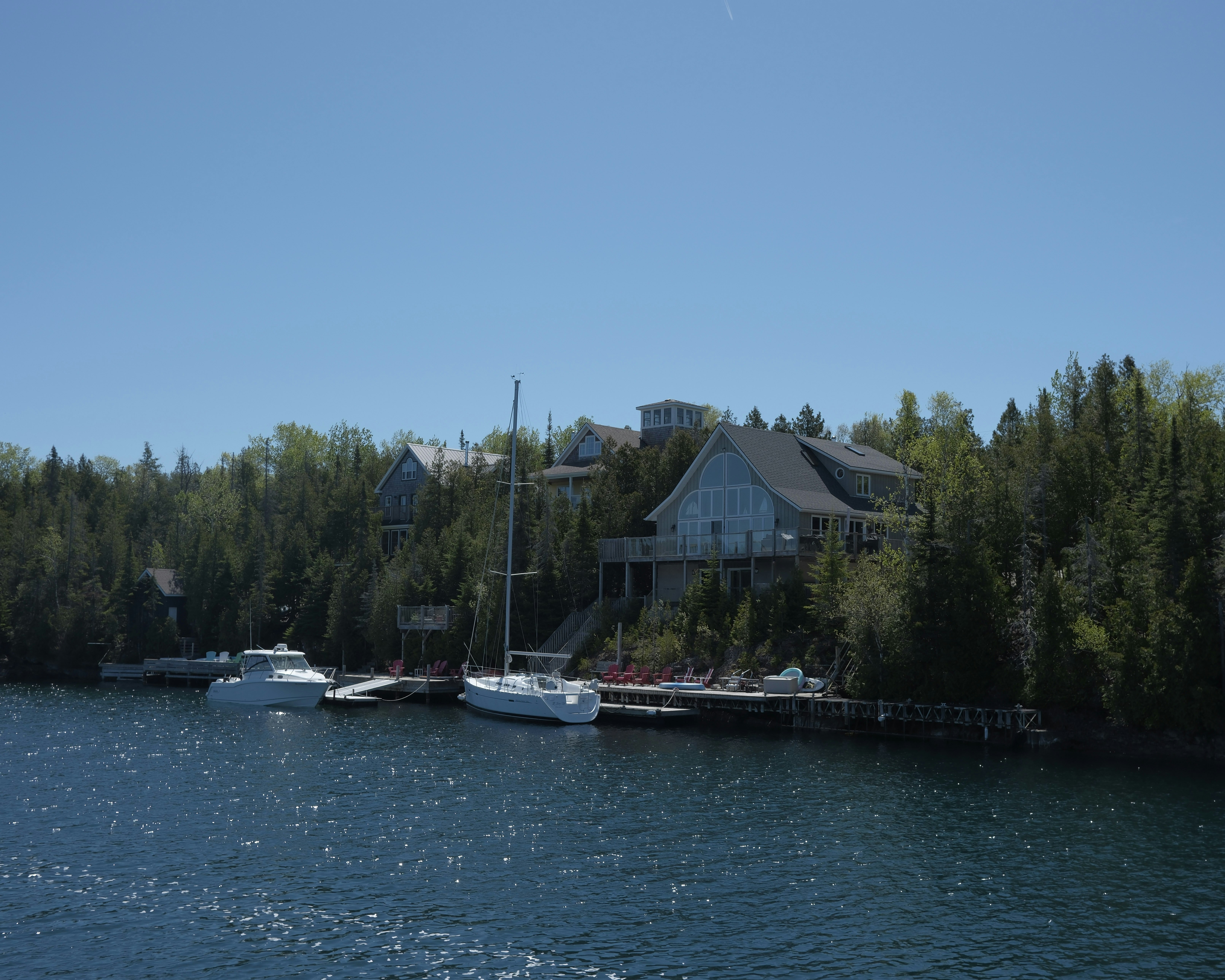 A low-angle shot showing luxury lakefront houses nestled into a thick forest of tall green pine trees. A white speedboat and a large sailboat are tied to the private wooden docks along the edge of the deep blue water. The sun makes the water sparkle beautifully in the foreground. This image highlights an upscale, relaxing lakeside lifestyle, making it a great visual asset for real estate agencies, luxury lifestyle magazines, or high-end apparel sponsors.