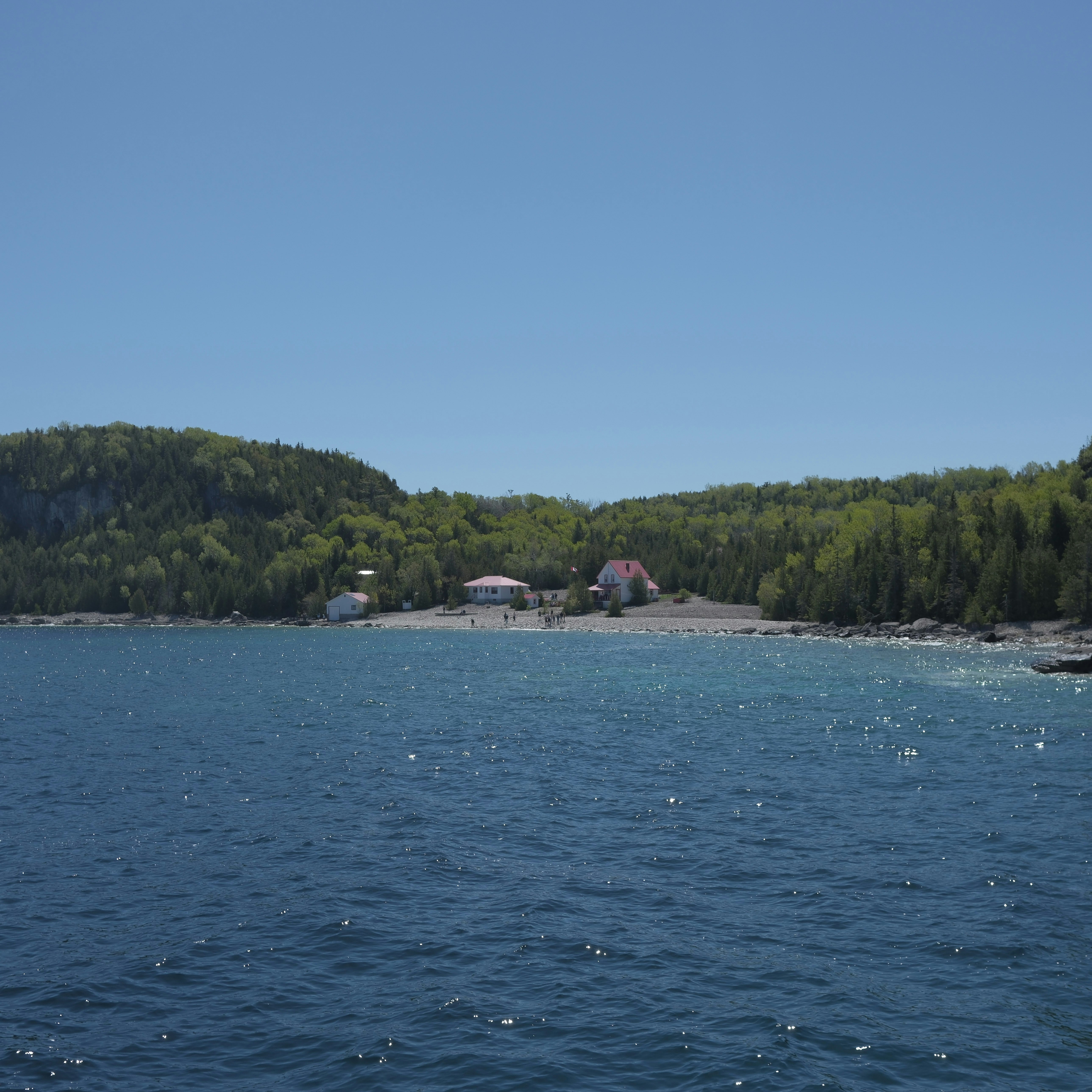 A wide horizontal landscape tracking a peaceful pebble beach cove with crystal-clear turquoise water. In the background, white and red cottage houses sit along the shore, backed by a big hill covered in thick green trees under a soft blue sky. The camera captures the beautiful, tropical-looking colors of the fresh water perfectly with zero edits. This relaxing scene is a wonderful match for hotel sponsors, destination marketing boards, or family travel channels.