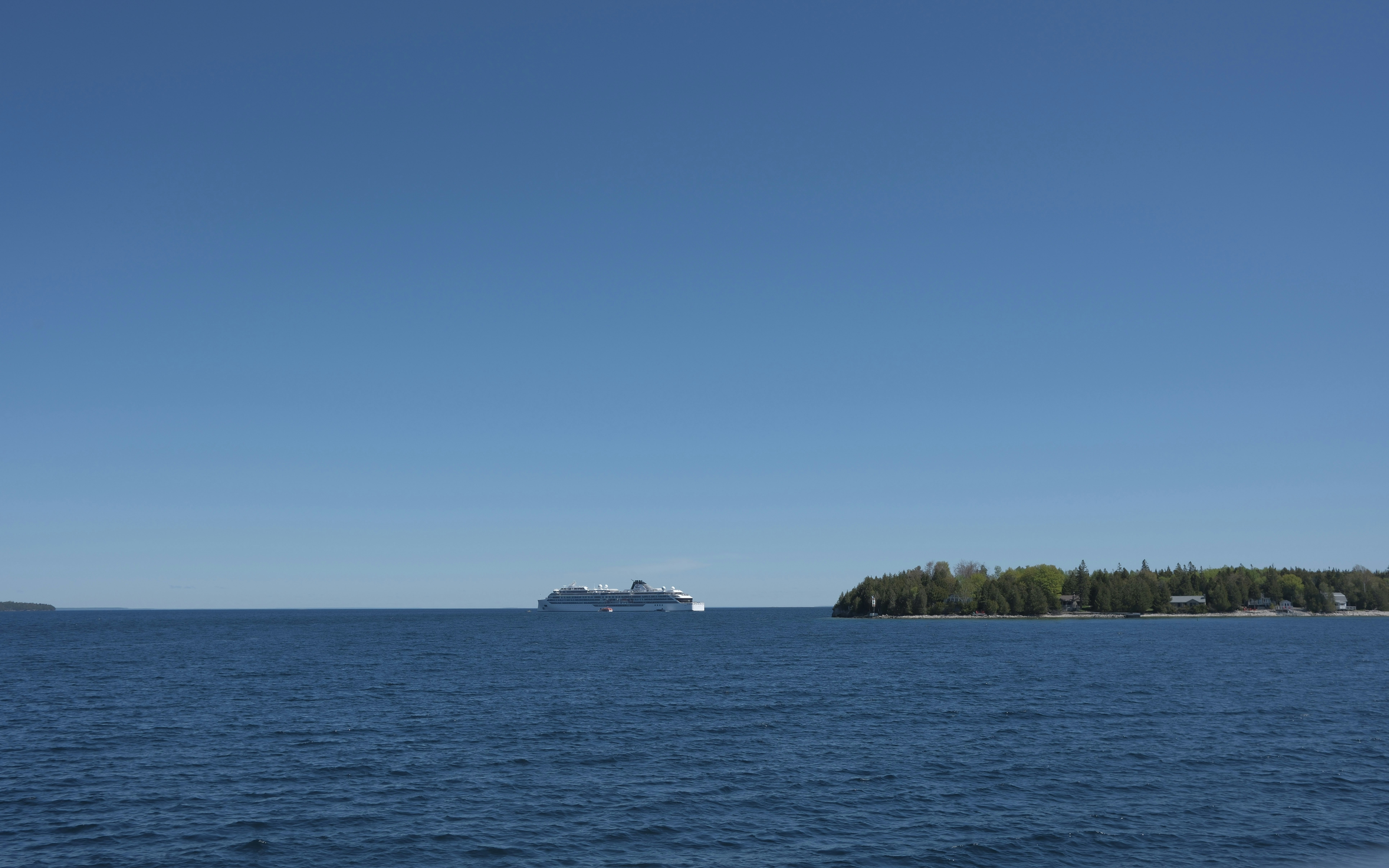A simple and peaceful view of a big cruise ship far away on the calm blue horizon. On the right side, there is a small island covered in dense green trees under a huge, cloudless blue sky. I love how clean and open this shot is, leaving lots of empty space for text or titles. This high-quality raw frame is a great match for cruise line advertisements, travel channel promotions, or international leisure magazines looking for serene ocean art.
