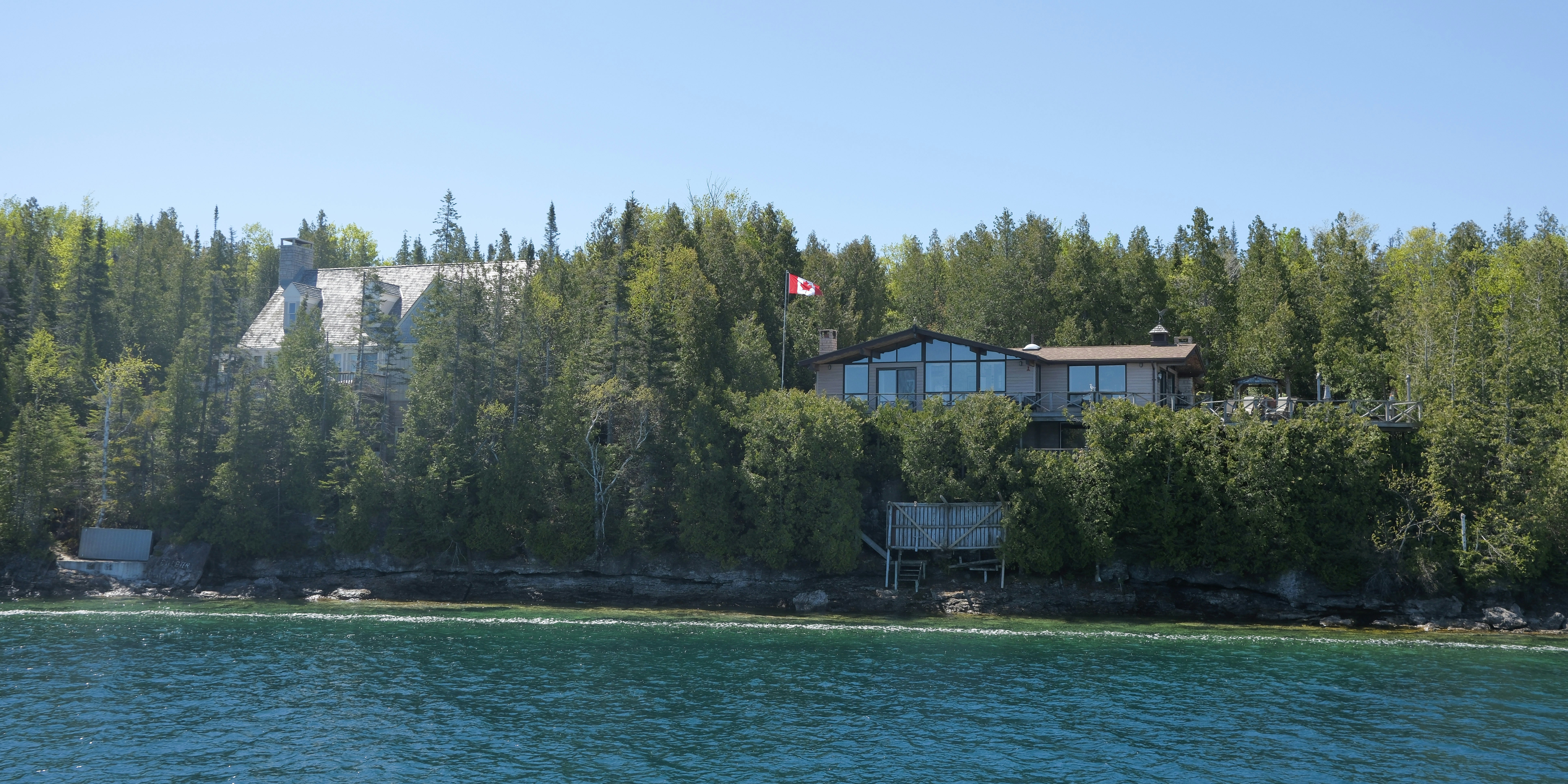 A beautiful view of two modern cottage homes built right on a rocky cliffside by the water. A Canadian flag waves proudly from a pole in the middle of the dense green forest surrounding the properties. The emerald-colored lake water in the foreground looks incredibly clear and inviting. This unedited photo shows a perfect balance of country living and beautiful nature, which is exactly what lifestyle brands and travel blogs look for.