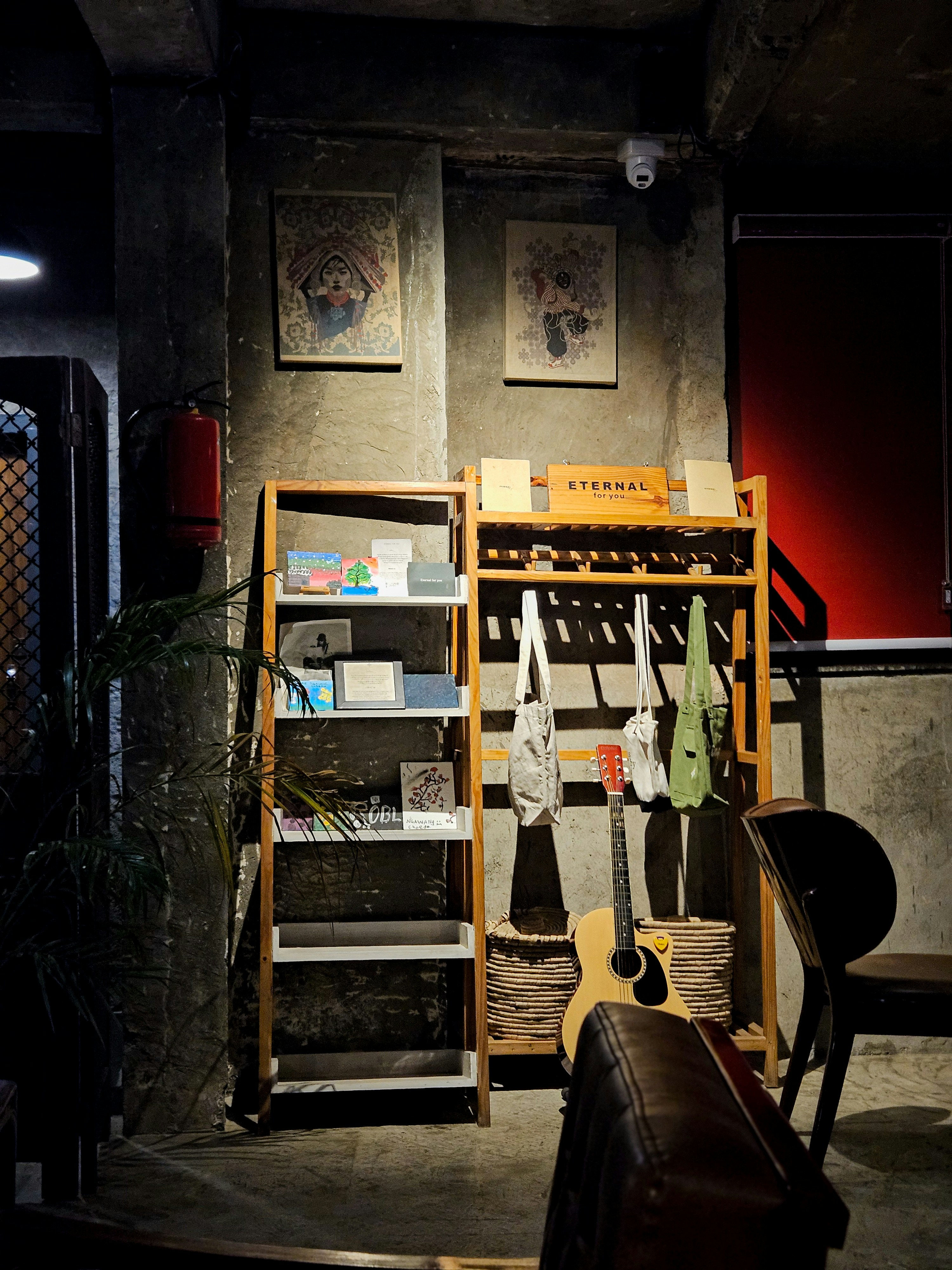 A cozy, industrial-styled interior scene featuring a wooden shelving unit with hanging bags and an acoustic guitar, set against a concrete wall with framed art. A woven basket and lounge seating complete the composition.