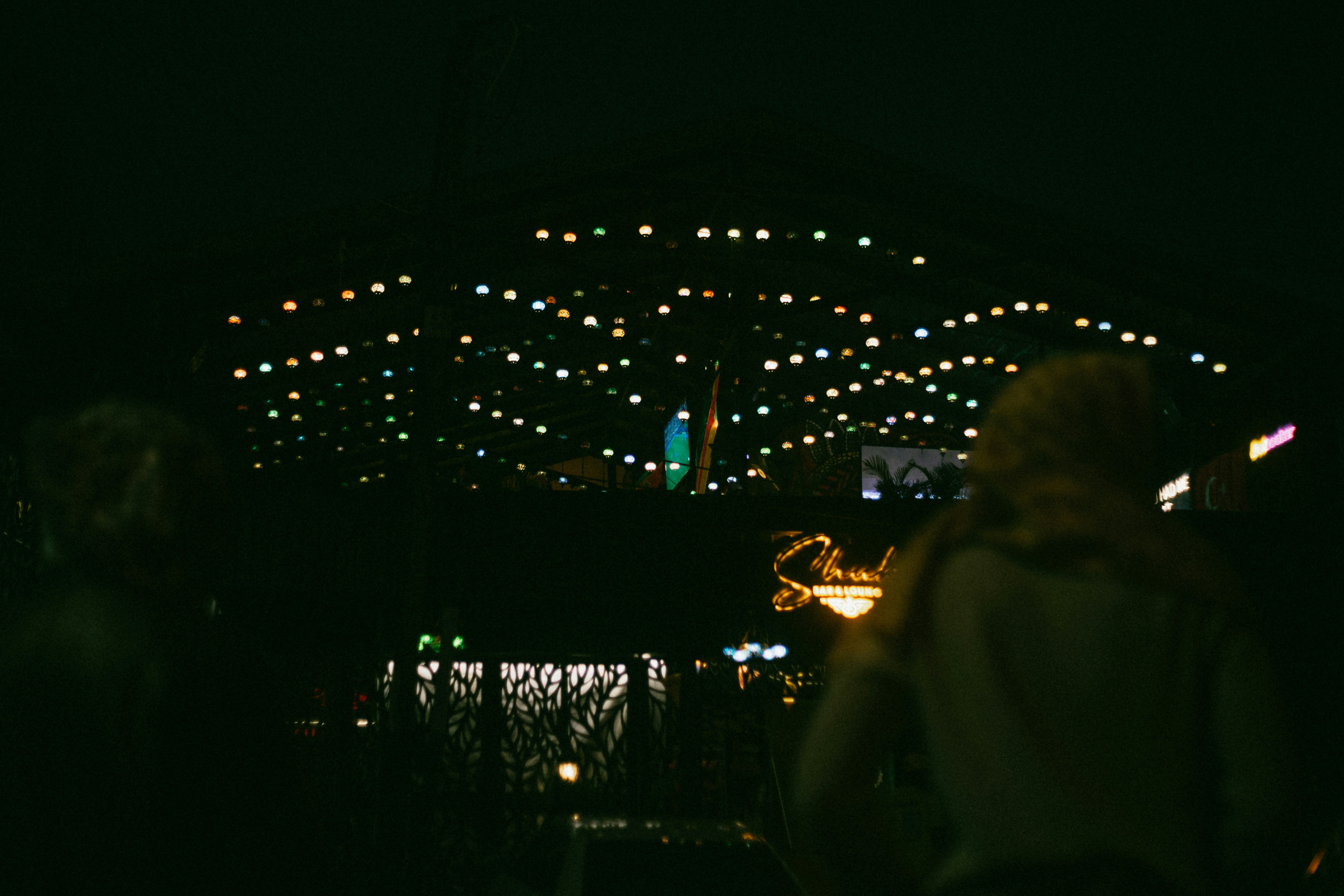 Dimly lit scene of a nighttime outdoor space with string lights and blurred figures in the foreground, suggesting a social gathering near a harbor or pier; a logo-like sign glows in the background.