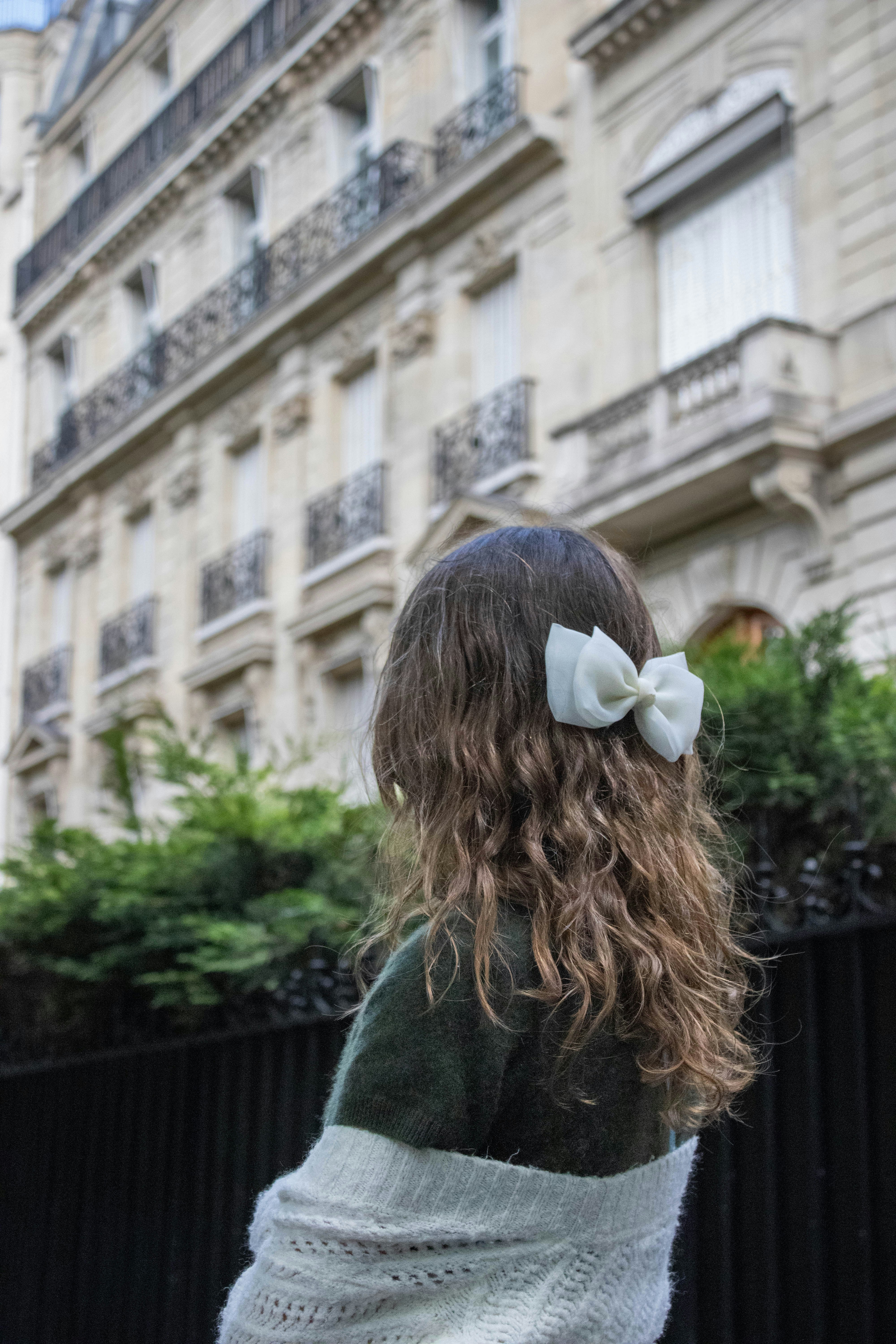 A girl with wavy hair tied by a pale bow stands with her back to the camera against a Parisian building facade; a knitted shawl drapes over her shoulders. This is a candid street portrait capturing texture and mood.