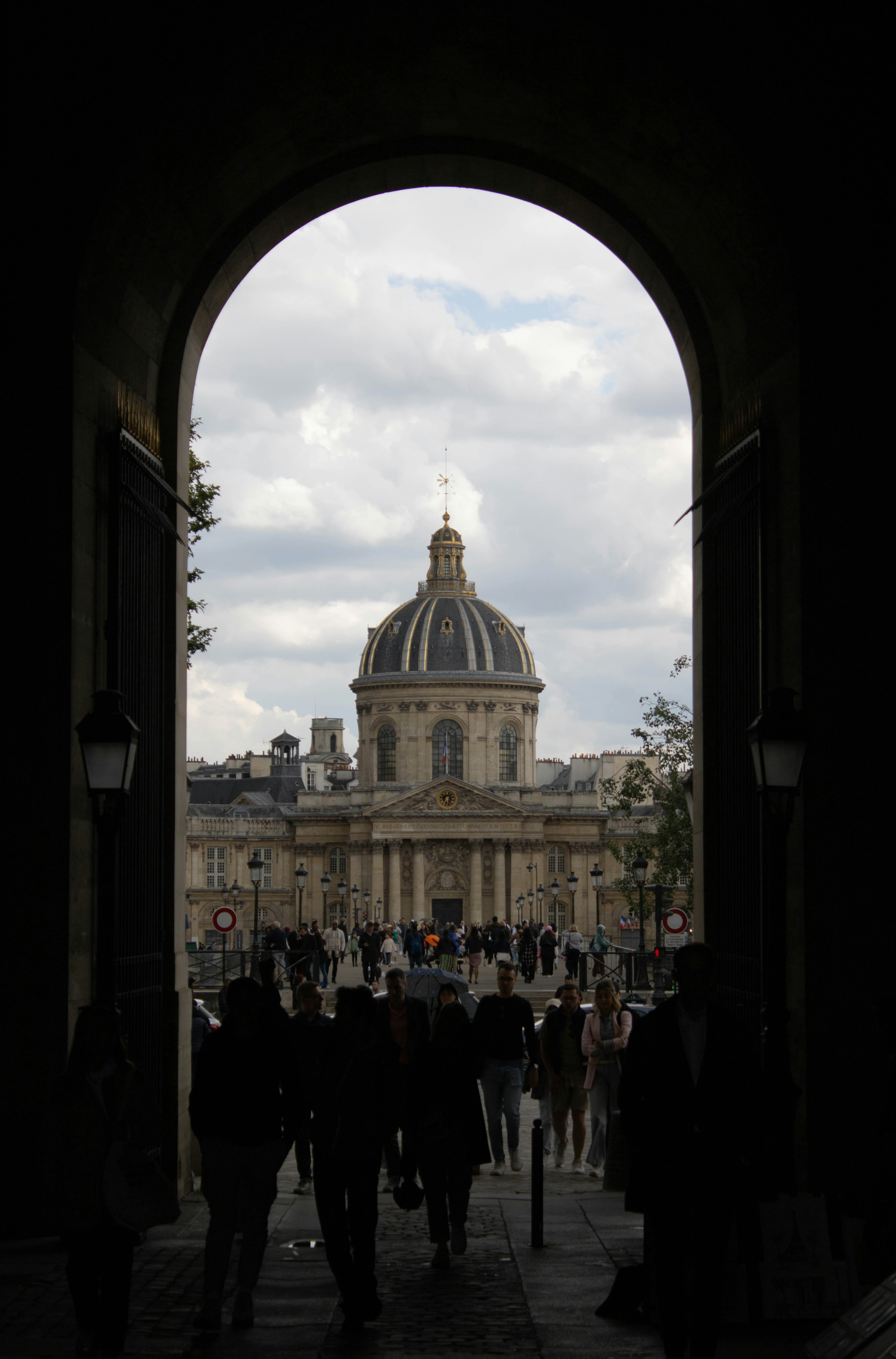 Dramatic view through a dark arch toward a grand domed cathedral with a crowd in the foreground; a street-scale, urban scene highlighting architectural symmetry.