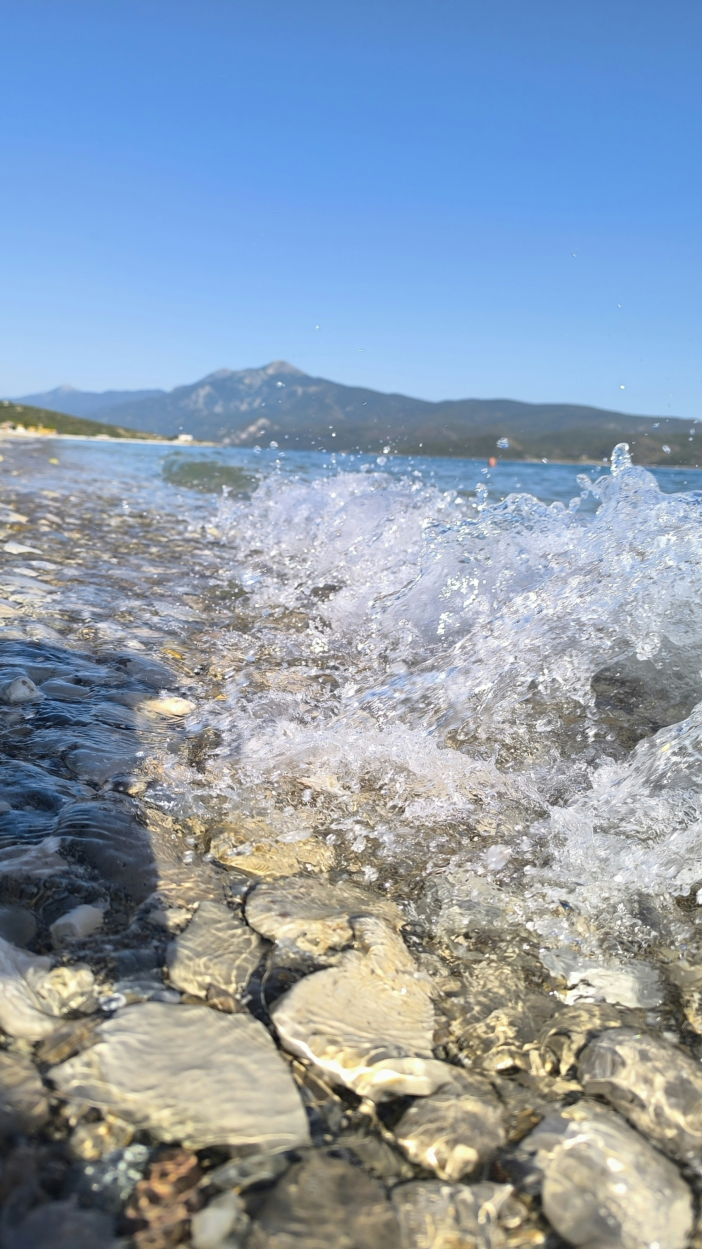 Close-up of curling waves washing over smooth pebbles along a rocky shoreline with distant mountains under a bright blue sky.