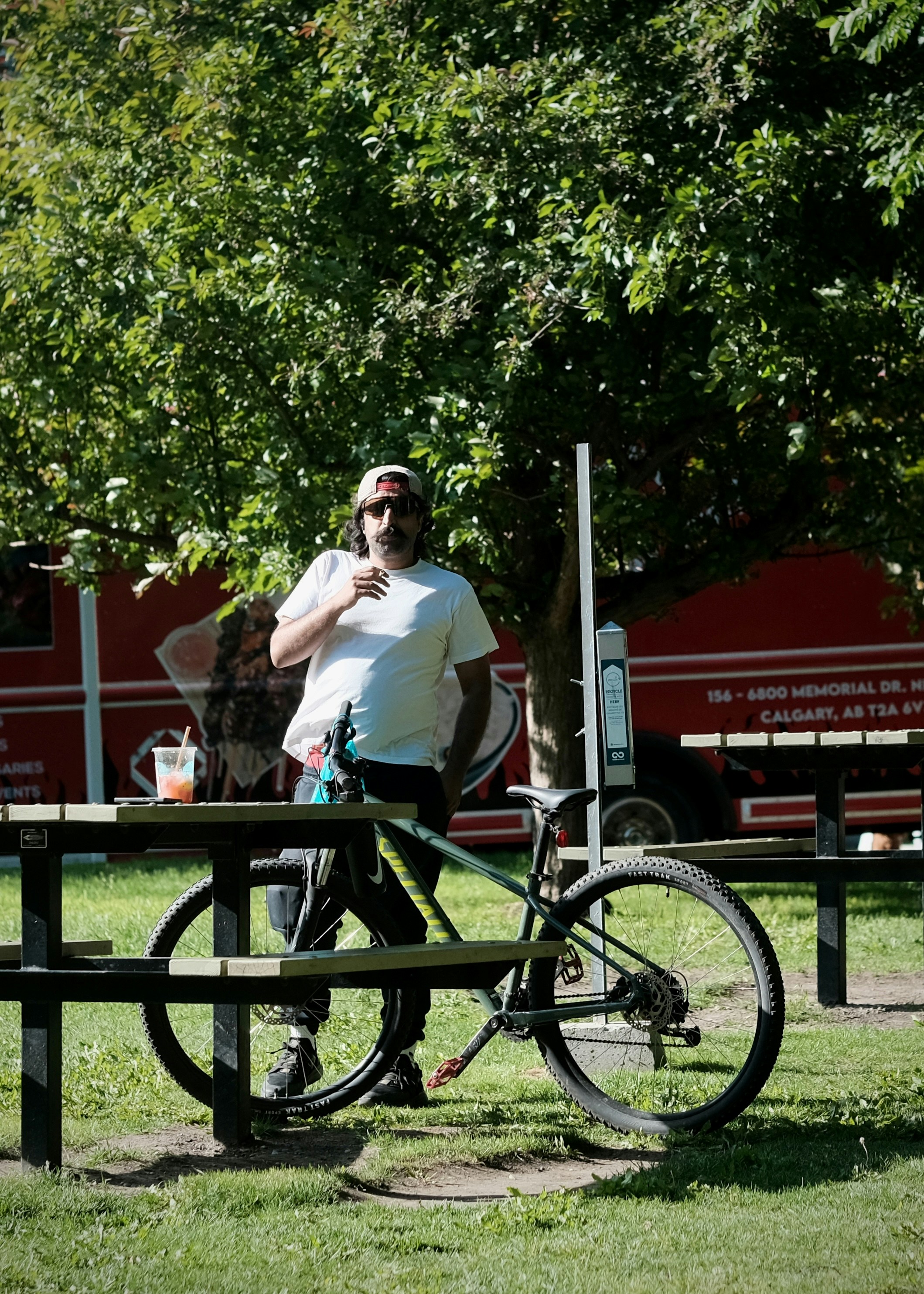 Man with white shirt leaning beside a bicycle at a park-side picnic table