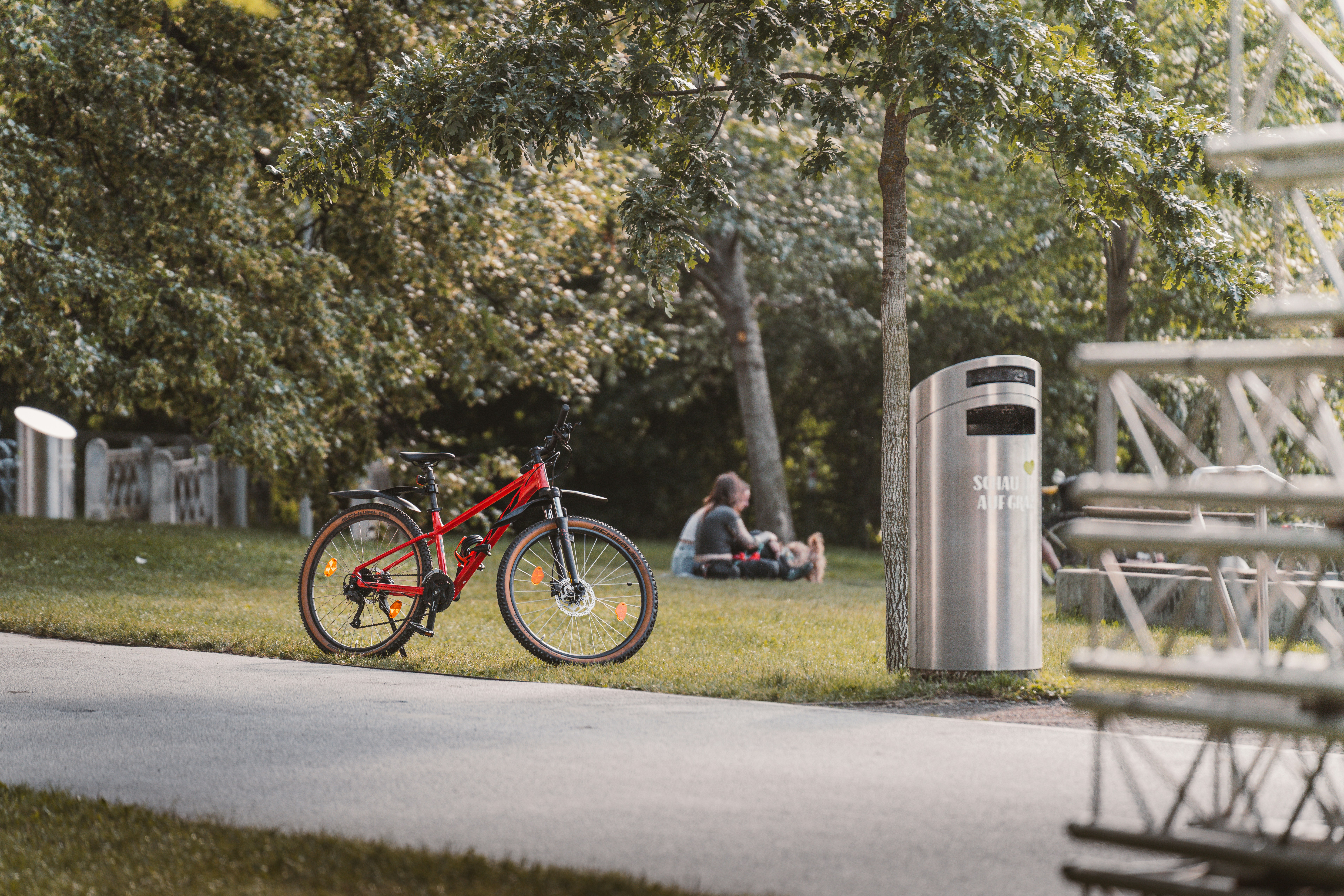 Red Bicycle at Park Edge with Gray Trash Can and Distant Portrait