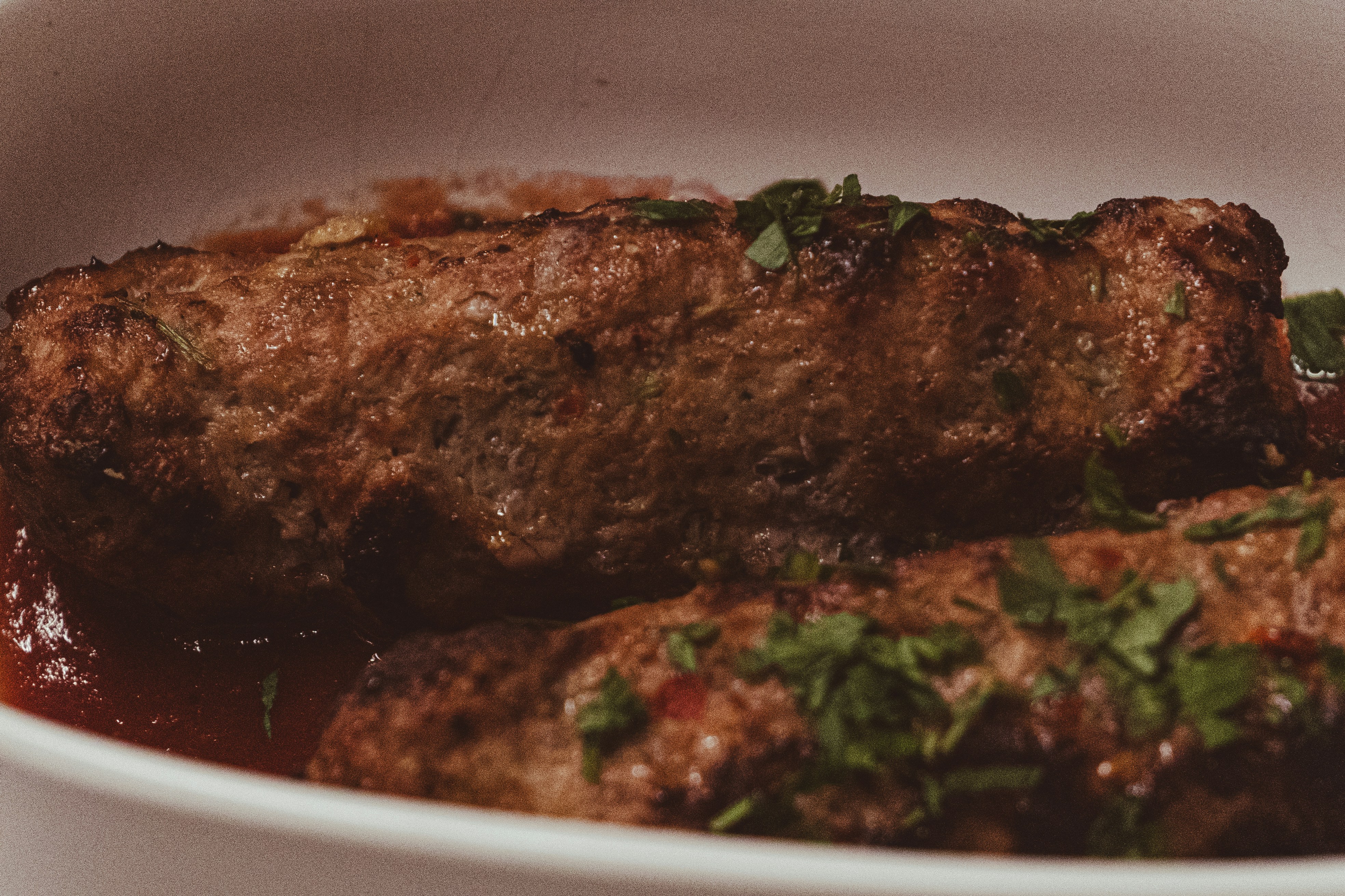Close-up of two browned meat patties simmering in a glossy tomato sauce, garnished with chopped parsley in a shallow white bowl.