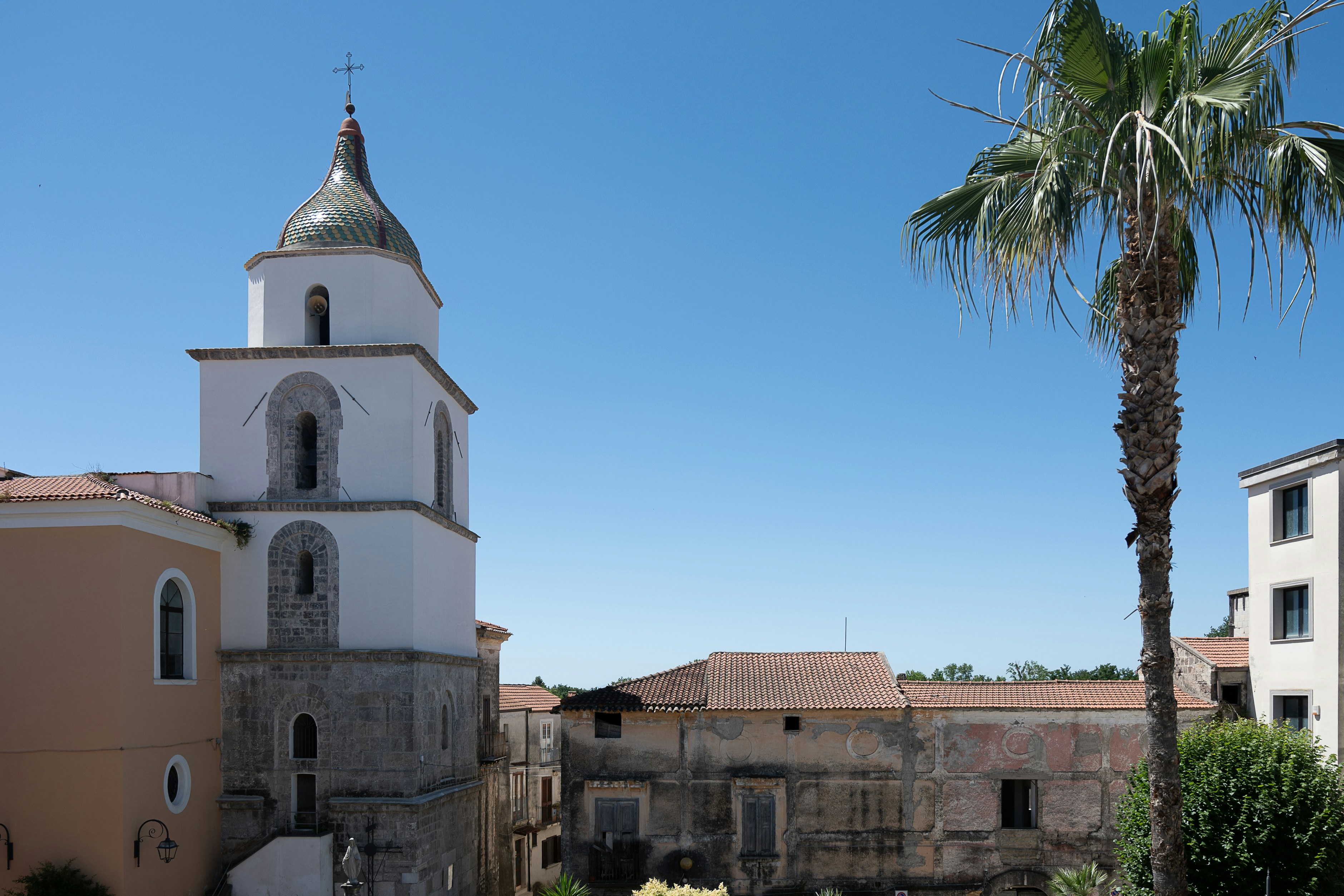 Sunlit white bell-tower structure bordered by weathered buildings and a lone palm, forming a quiet townscape.