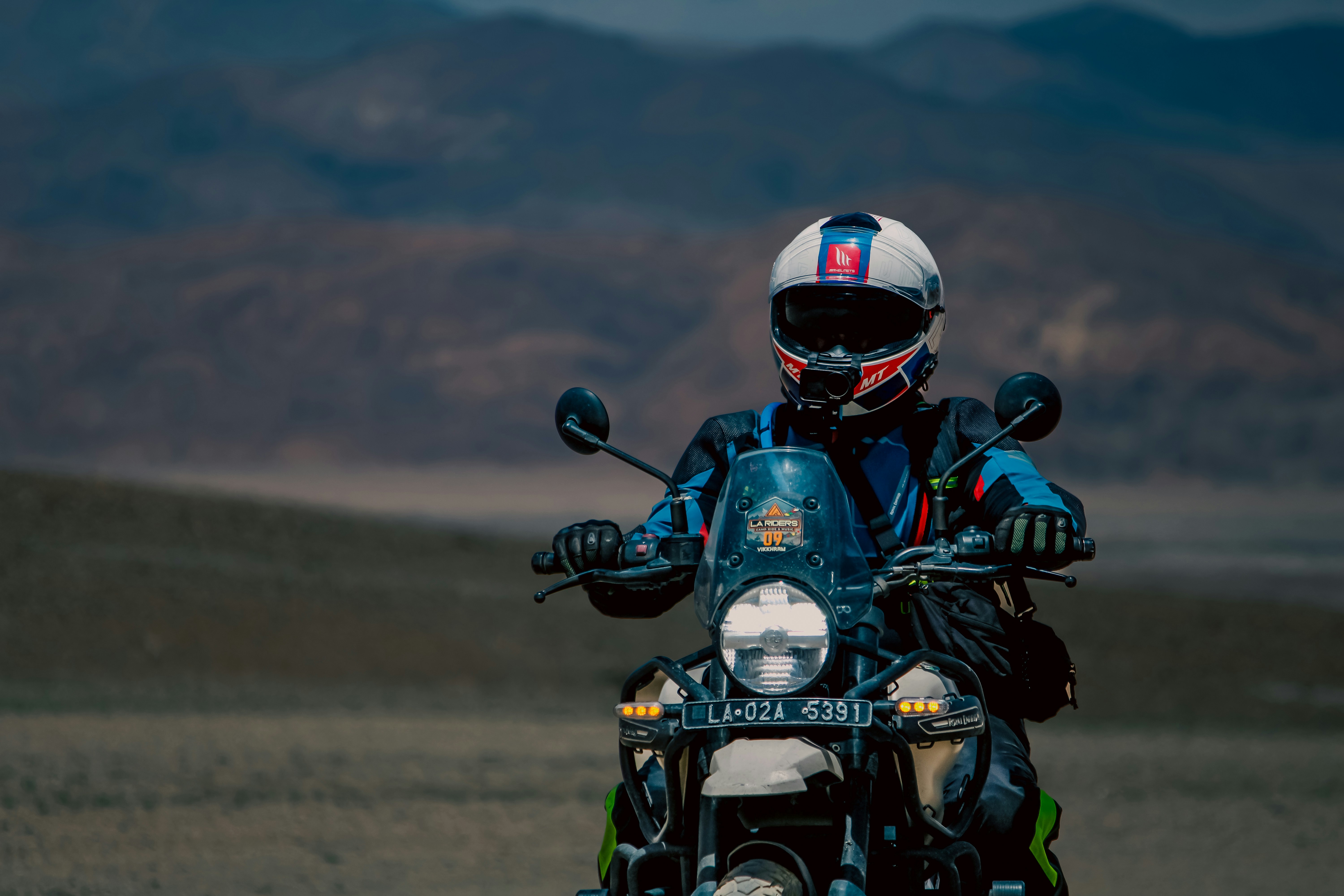 Motorcycle rider centered in a dusty landscape with mountains in the background; full face helmet and protective gear visible, headlight on, front-of-bike detail emphasized. Photograph captures action-ready posture and remote terrain.