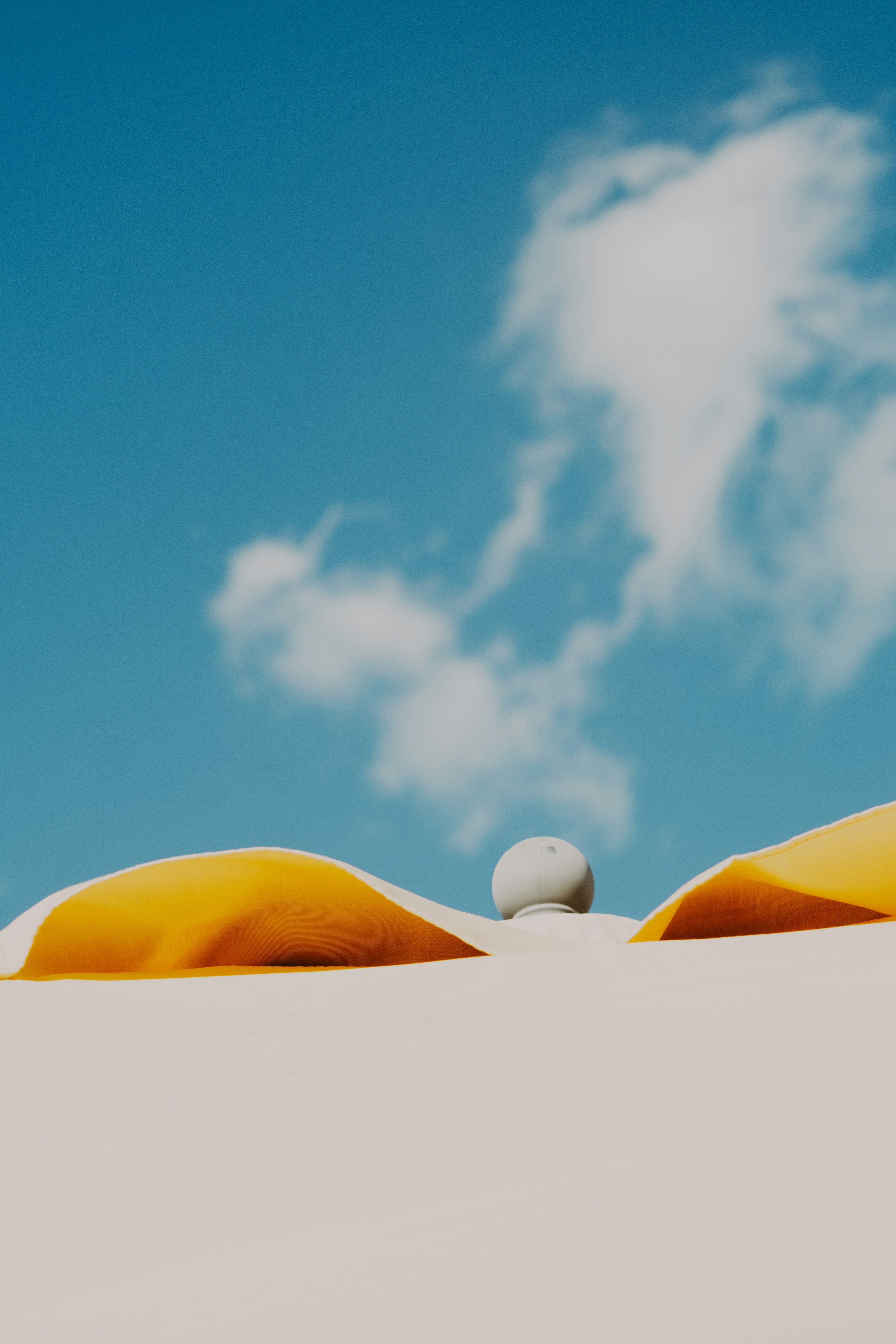 A minimal coastal dune scene with a bright blue sky and a white sand ridge, punctuated by a small rounded object at the horizon. This is a landscape photograph.