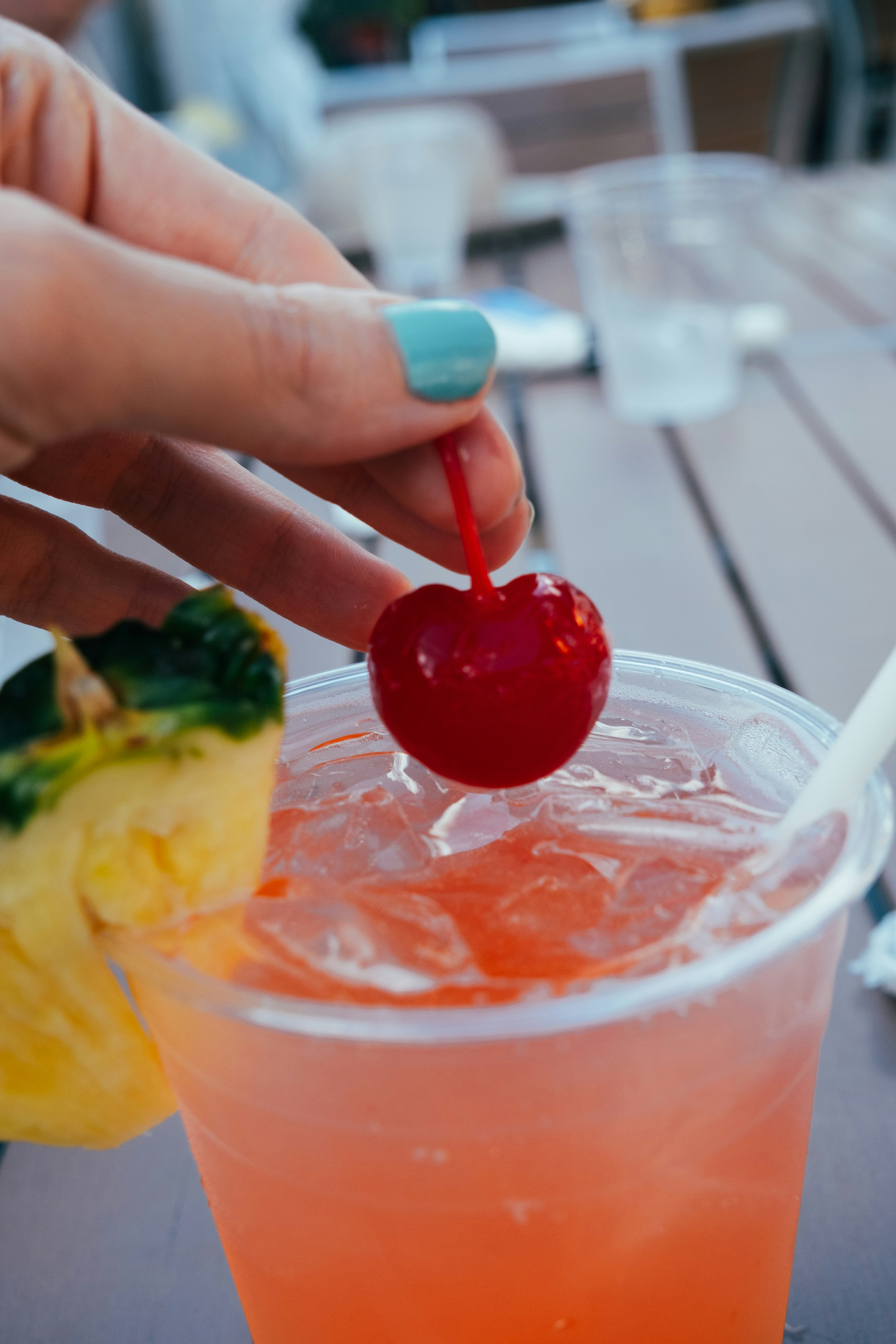 Bright red cherry held above a pink citrus beverage with ice, a pineapple garnish, and a blurred outdoor table scene in the background.