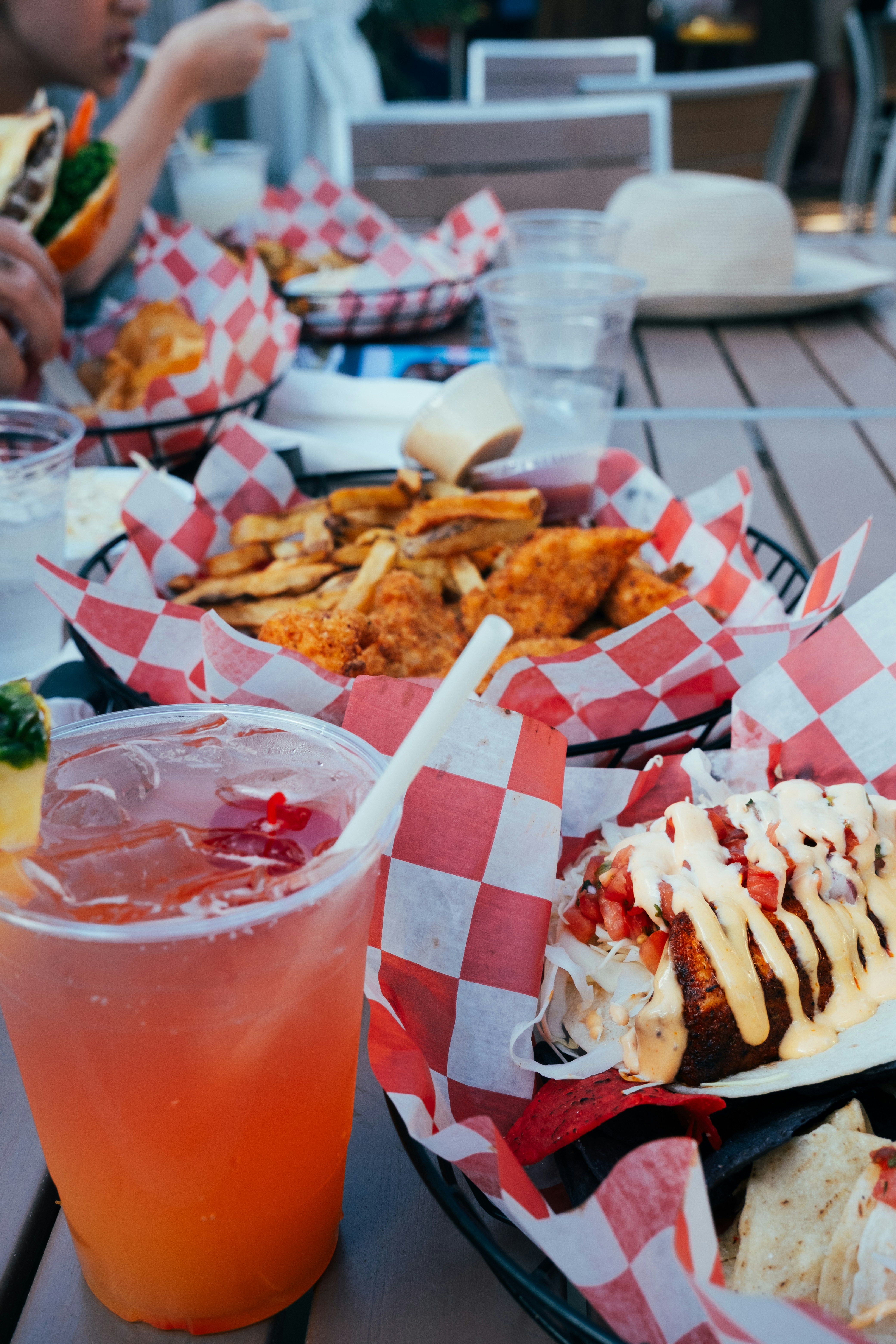 Plateful of fried chicken and chips with dipping sauce in a casual outdoor setting, foregrounded by a pink beverage; a dessert roll drizzled with sauce sits to the right, all lined with red-and-white checkered paper.
