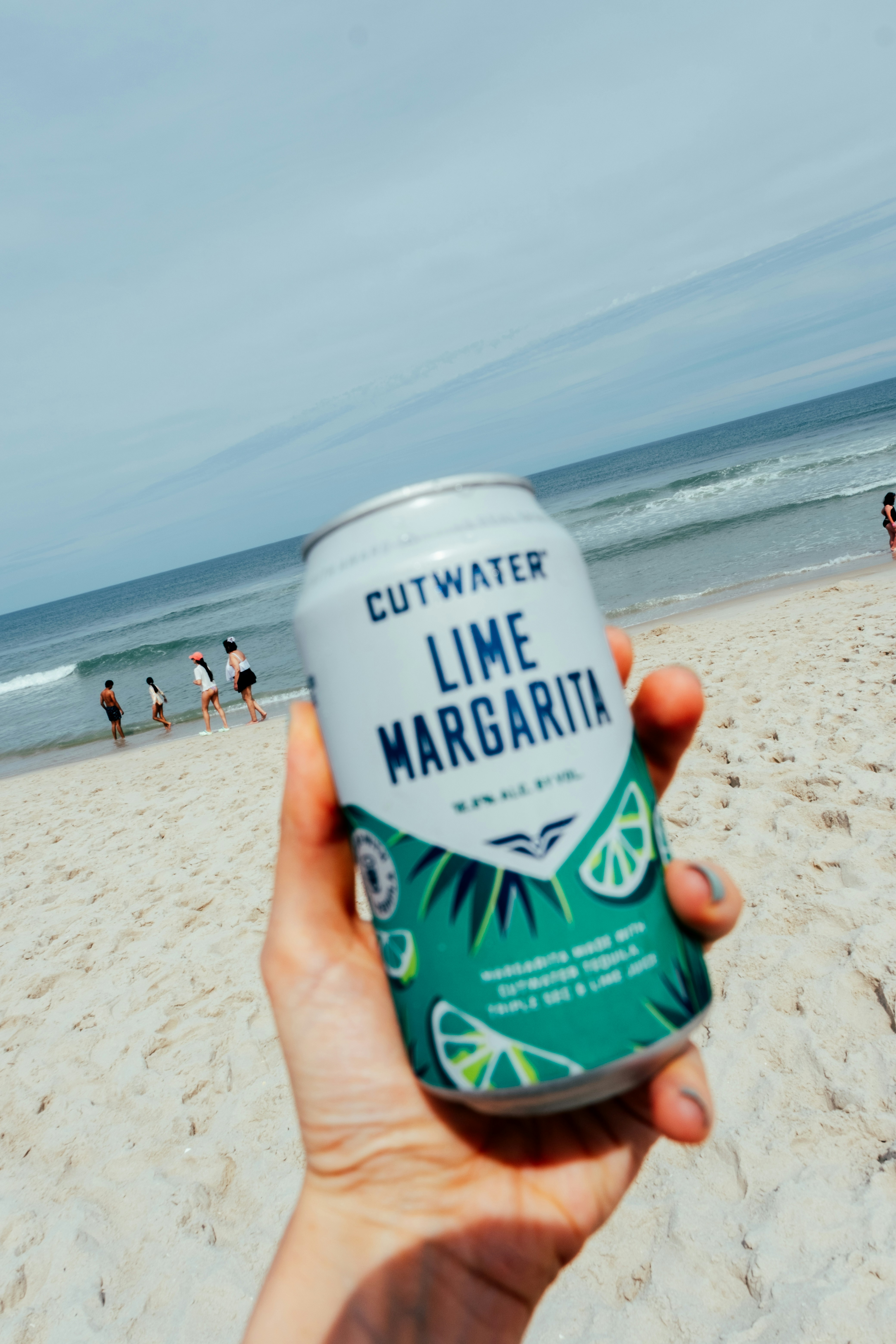 Lime Margarita in Focus: Beachside Hand and Can Framed by Surf