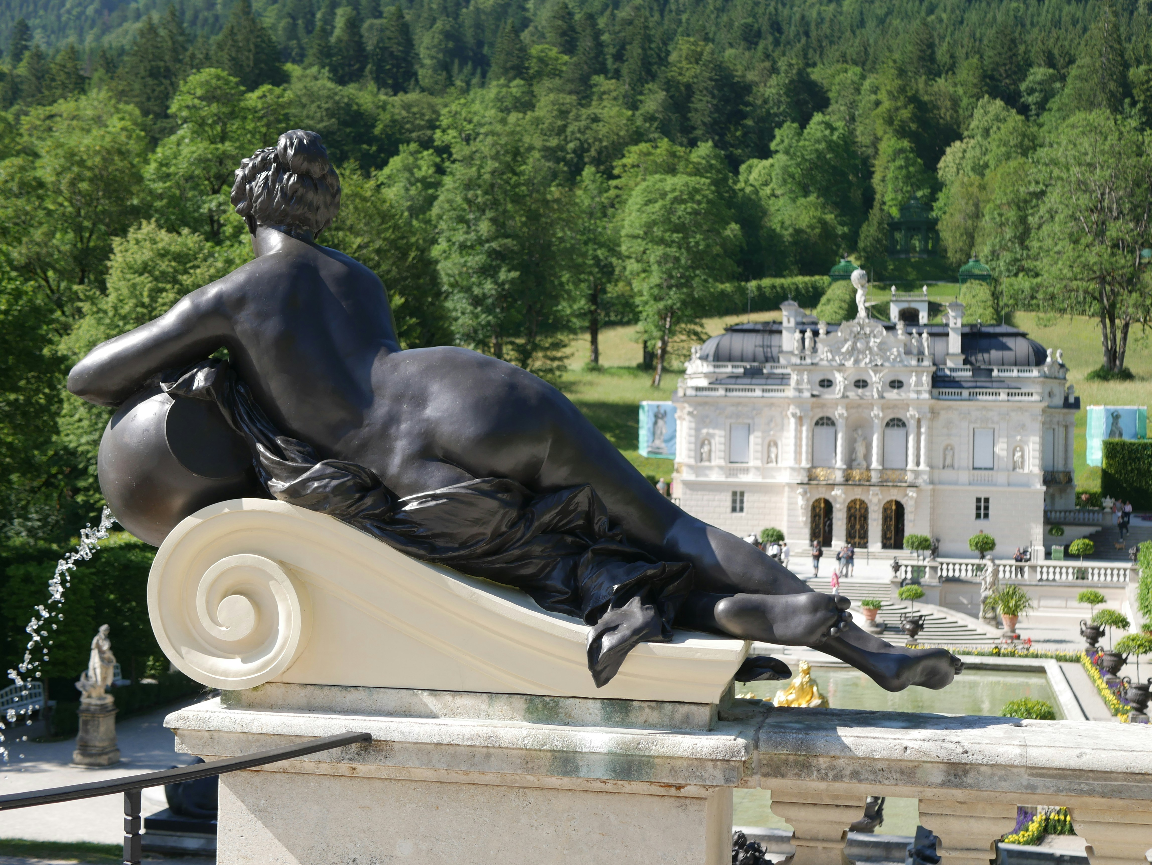 Bronze reclining sculpture atop a curved pedestal with a grand chateau and formal gardens in the background.
