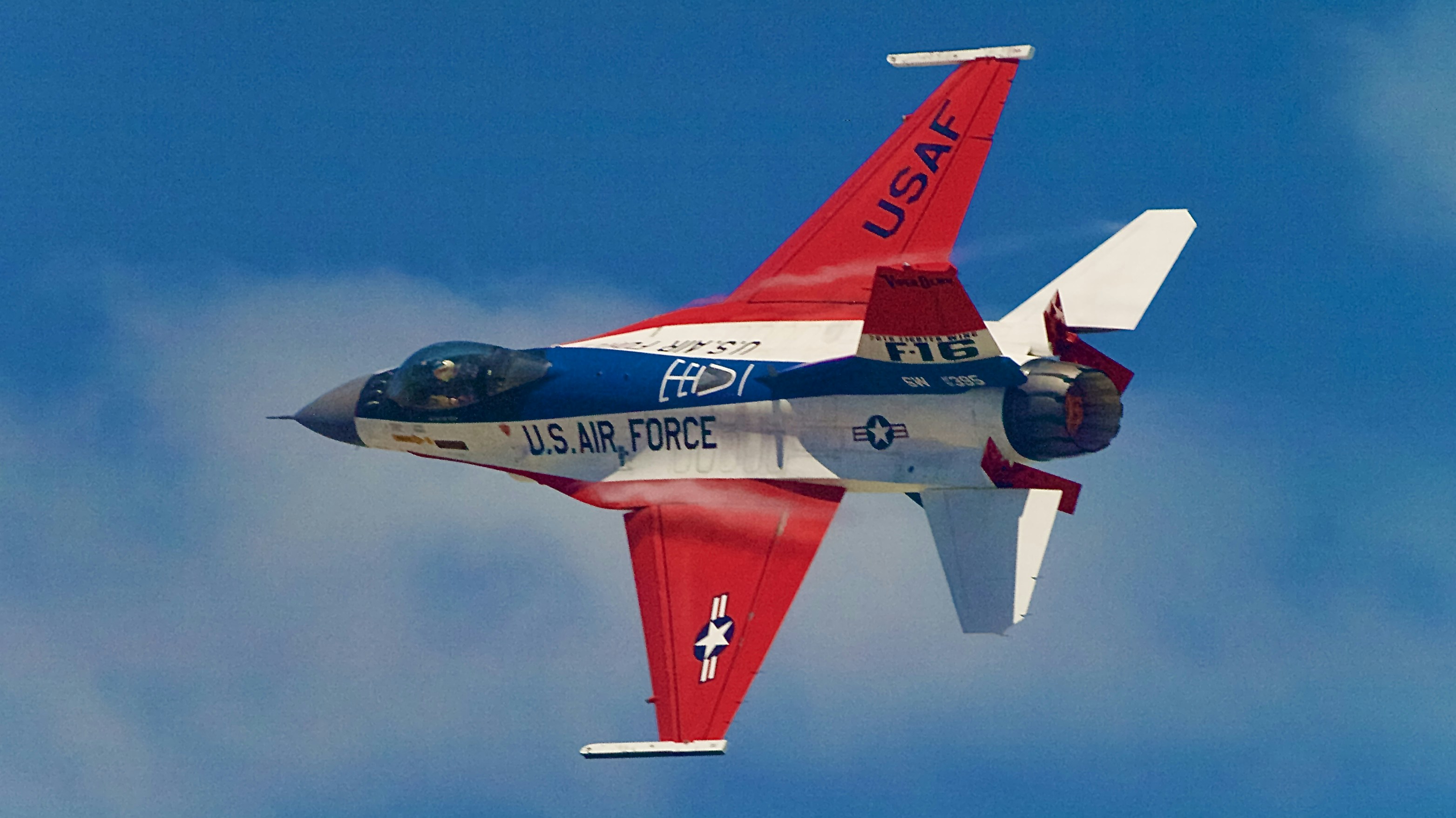 F-16 fighter jet soars against a bright blue sky with light clouds, displaying bold red vertical stabilizers and white nose markings.