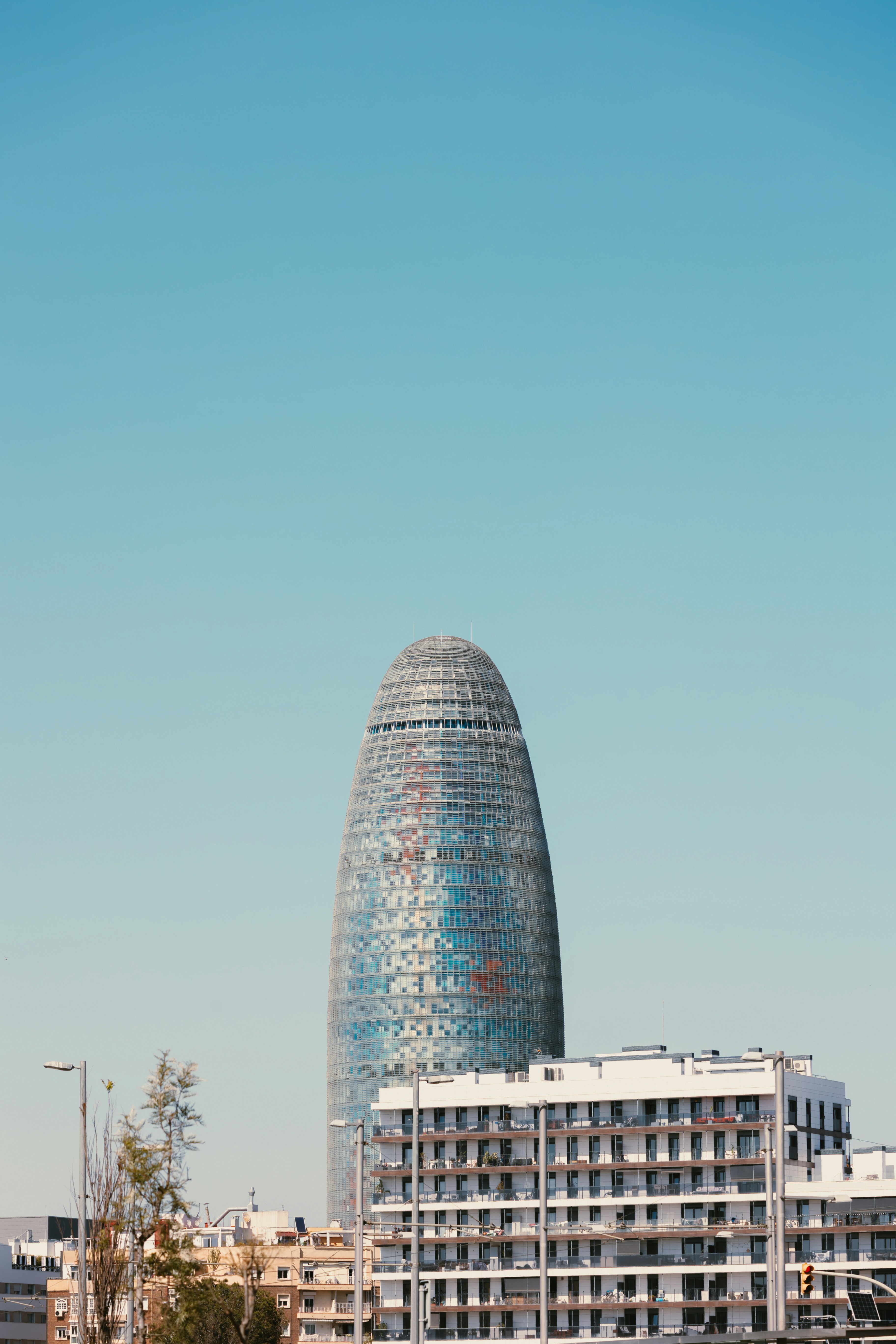Wide angle view of the glass-facade Torre Glòries skyscraper towering over a white apartment building against a clear blue sky in Barcelona.