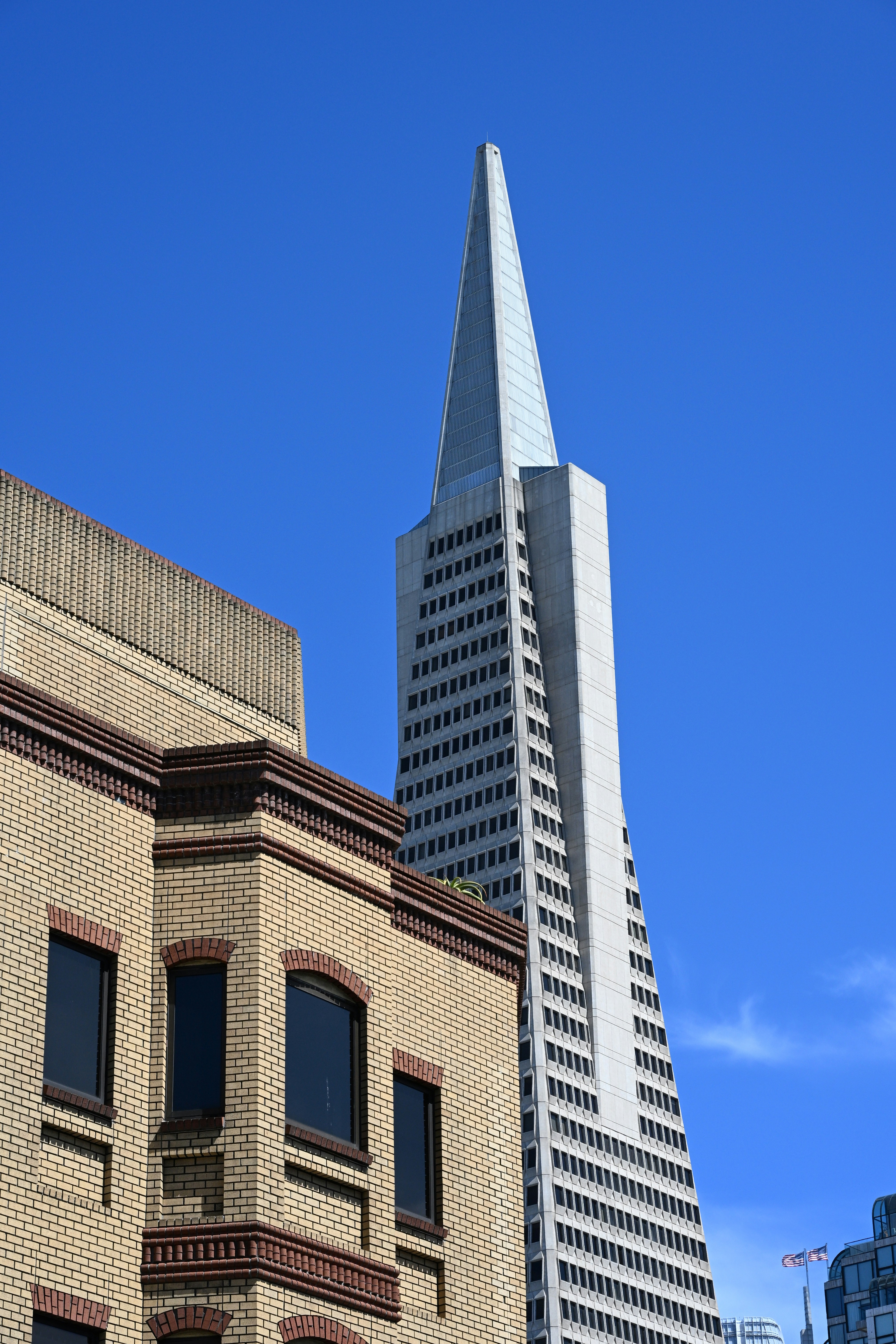 Tall, angular skyscraper rising beside a brick building under a crisp blue sky; the scene emphasizes architectural contrast and verticality in an urban landscape.