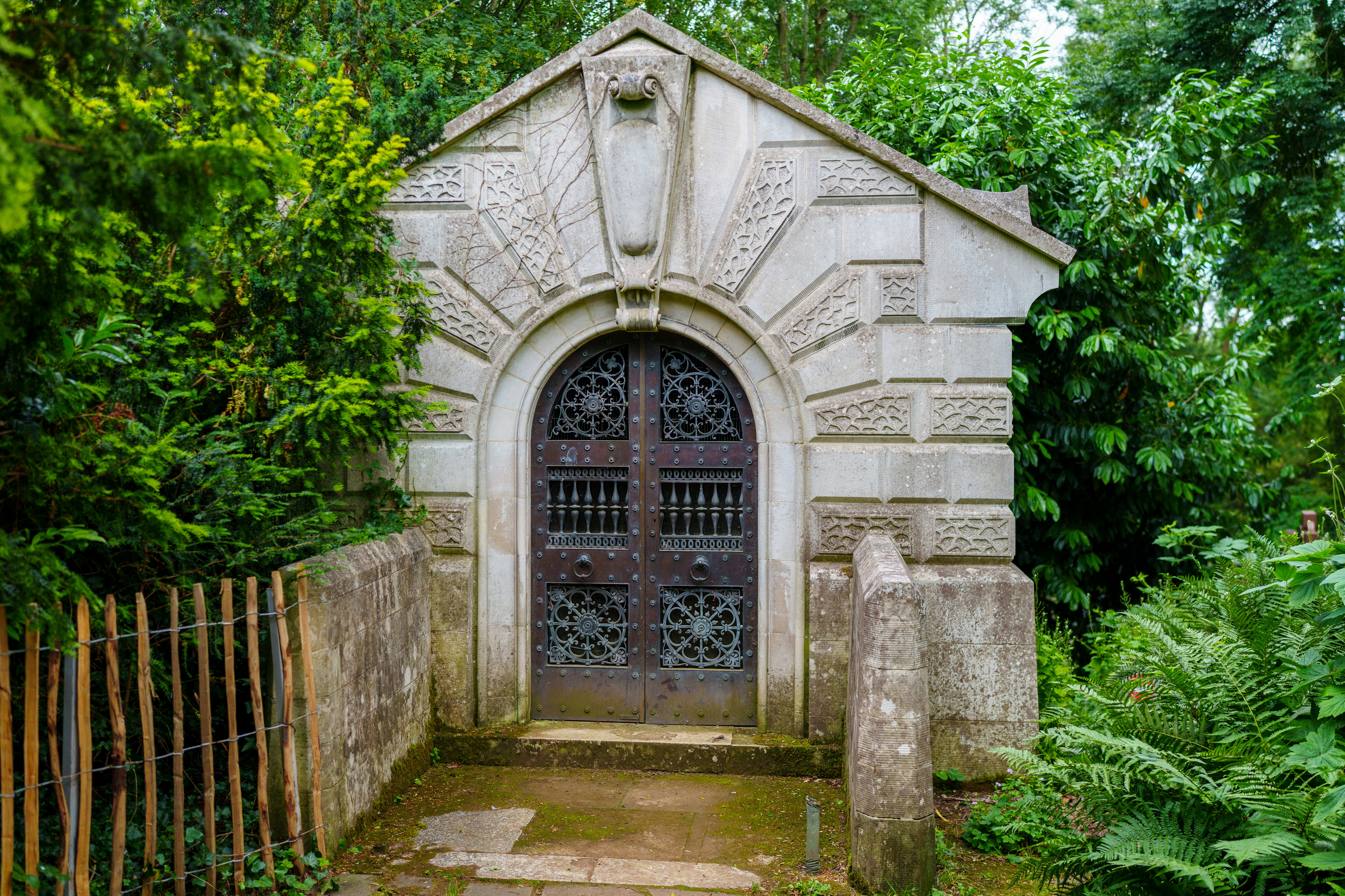 Ornately carved Victorian mausoleum with wrought-iron doors, surrounded by ferns and foliage