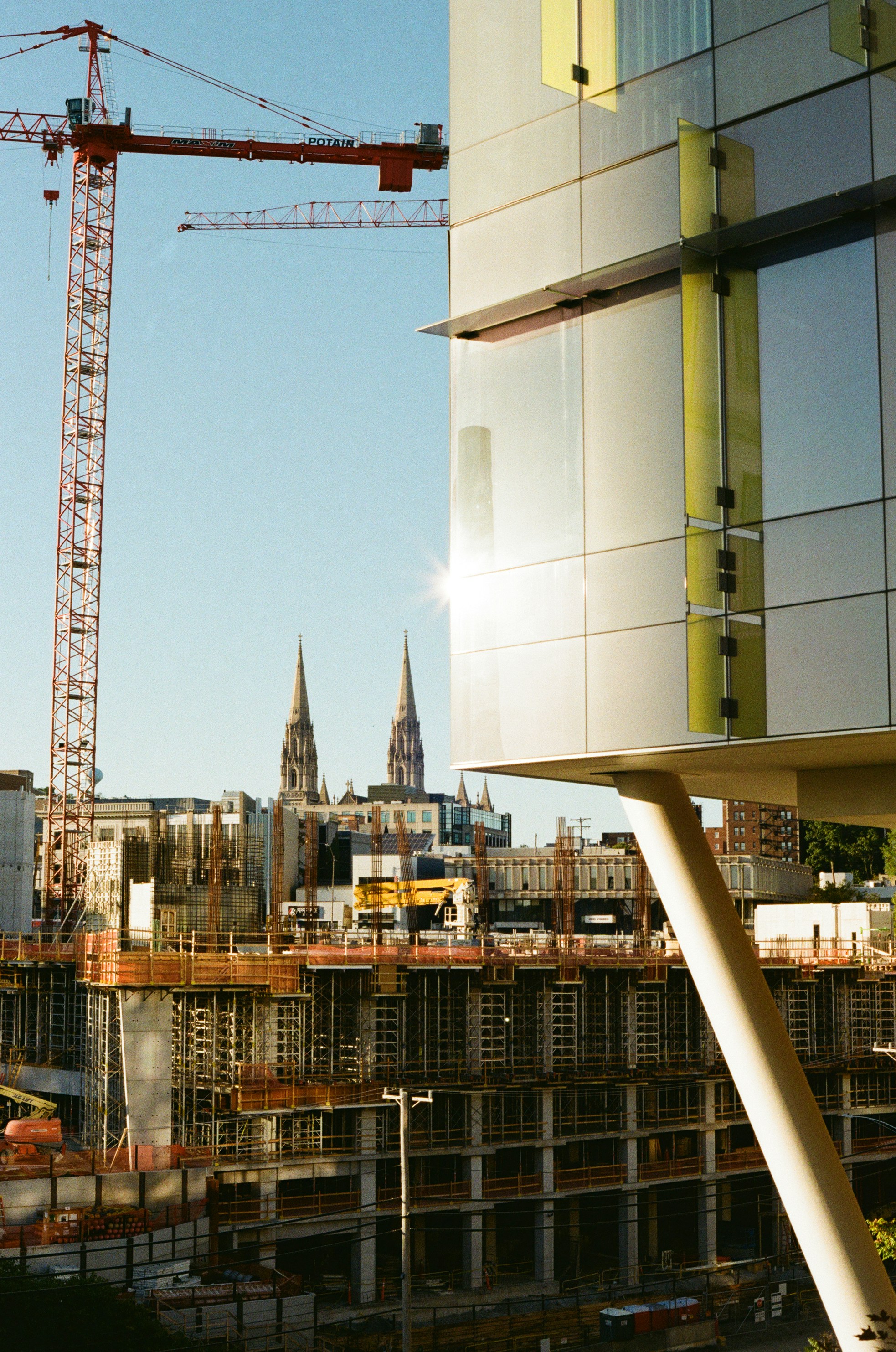 Construction site with cranes and building forms seen beneath a modern glass-clad structure against a clear sky.